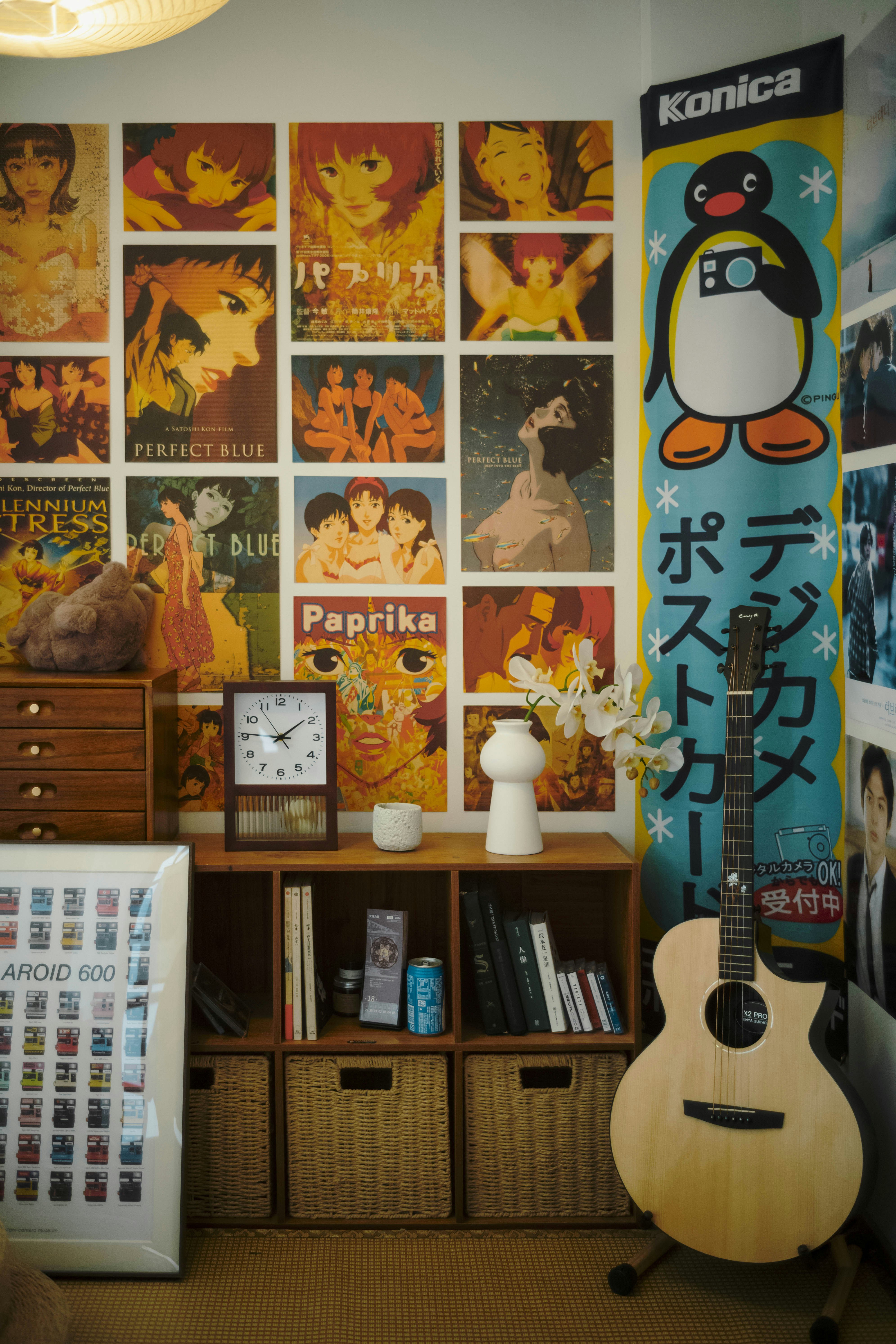 Cozy room with a wall collage of colorful poster art and a guitar leaning by a bright Konica banner. A wooden cabinet holds a clock and decor, grounding the scene.