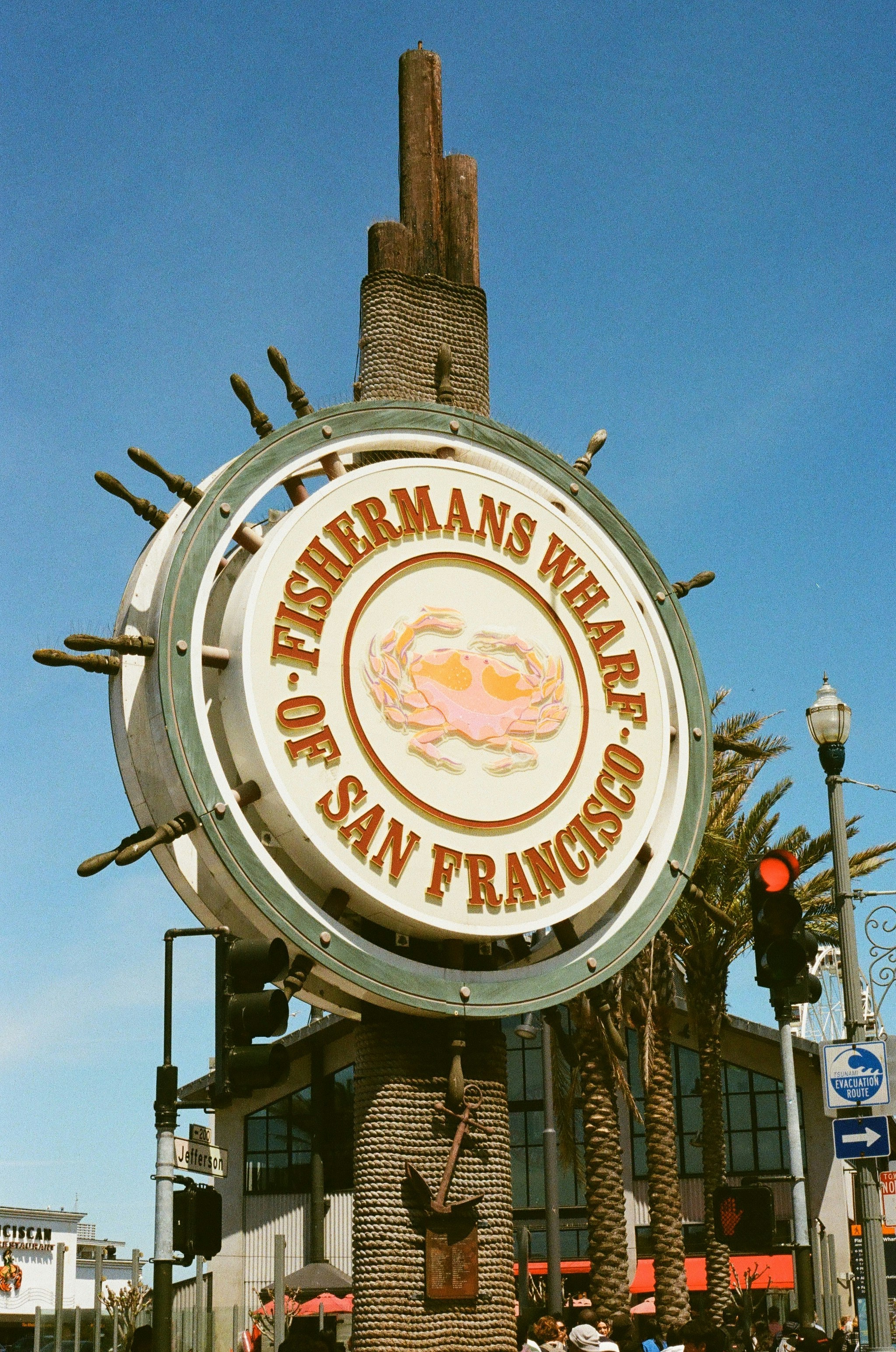 Large circular sign for Fisherman's Wharf, San Francisco, photographed against a clear blue sky. The emblem features rope detailing and bold lettering, marking a popular waterfront landmark.