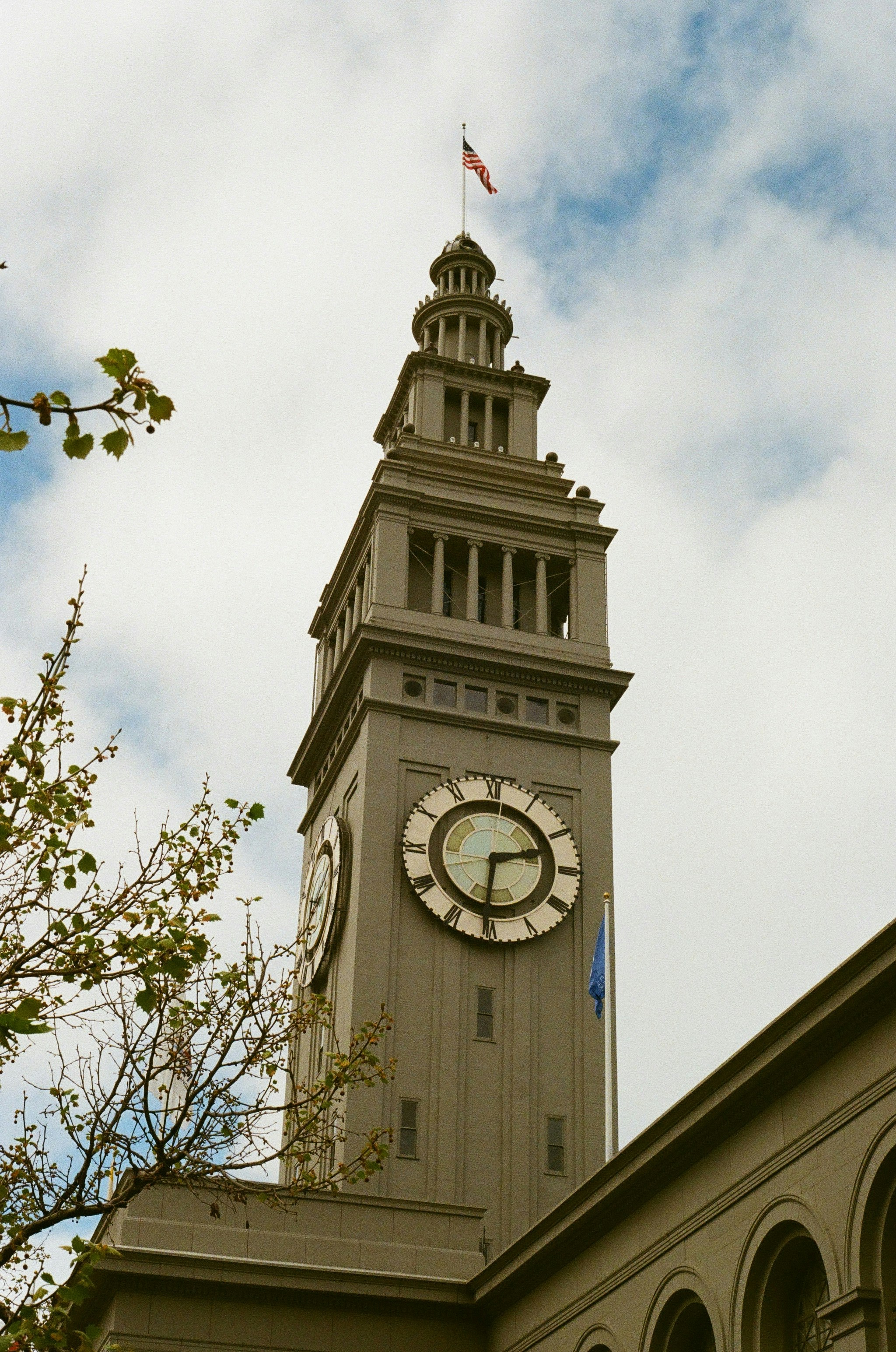 An imposing clock tower rises into a partly cloudy sky, topped by an American flag. Branches in the foreground frame the historic monument.