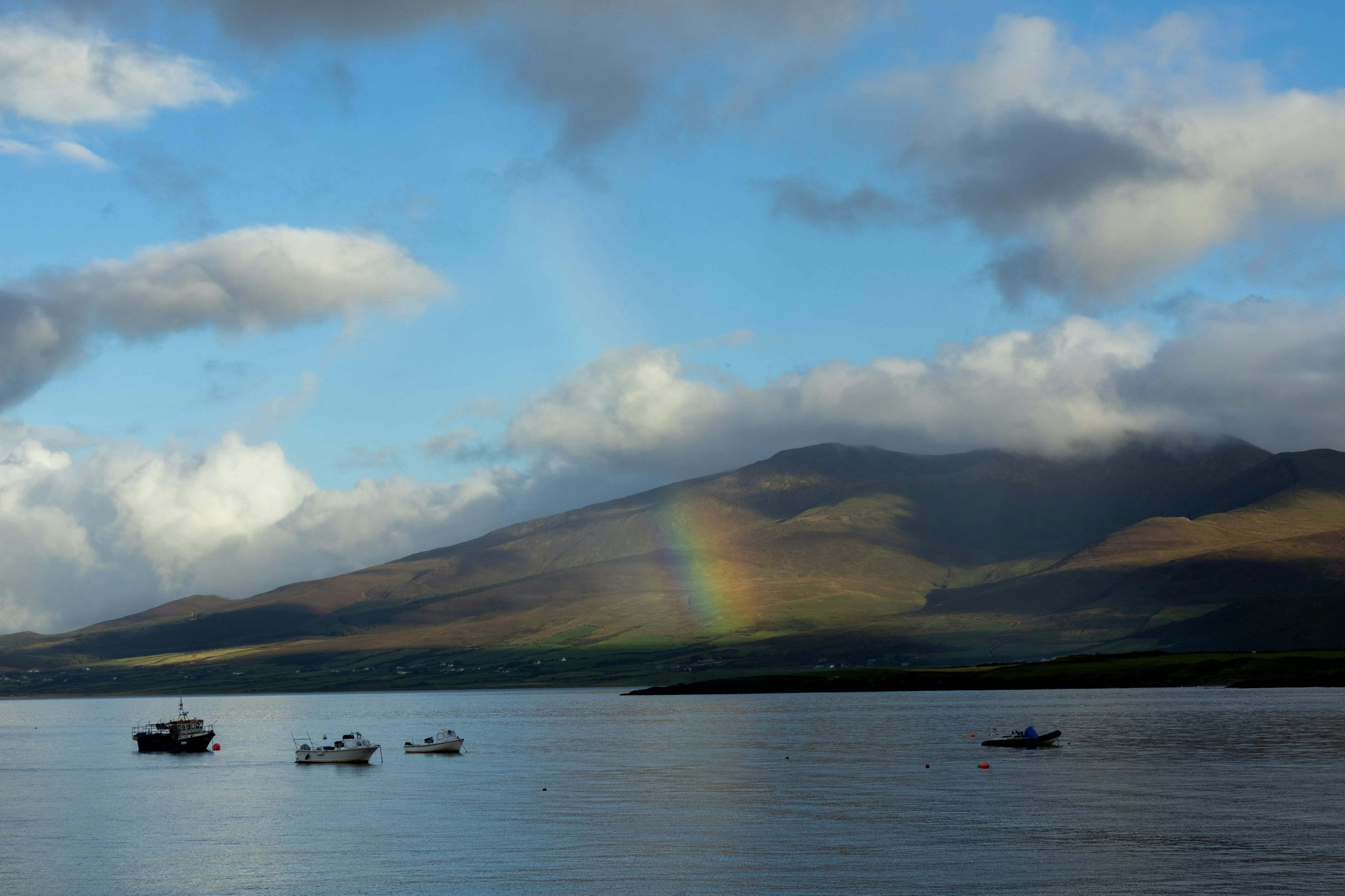 Boats floating on a calm lake with a rainbow arching over a distant mountain under a partly cloudy sky.