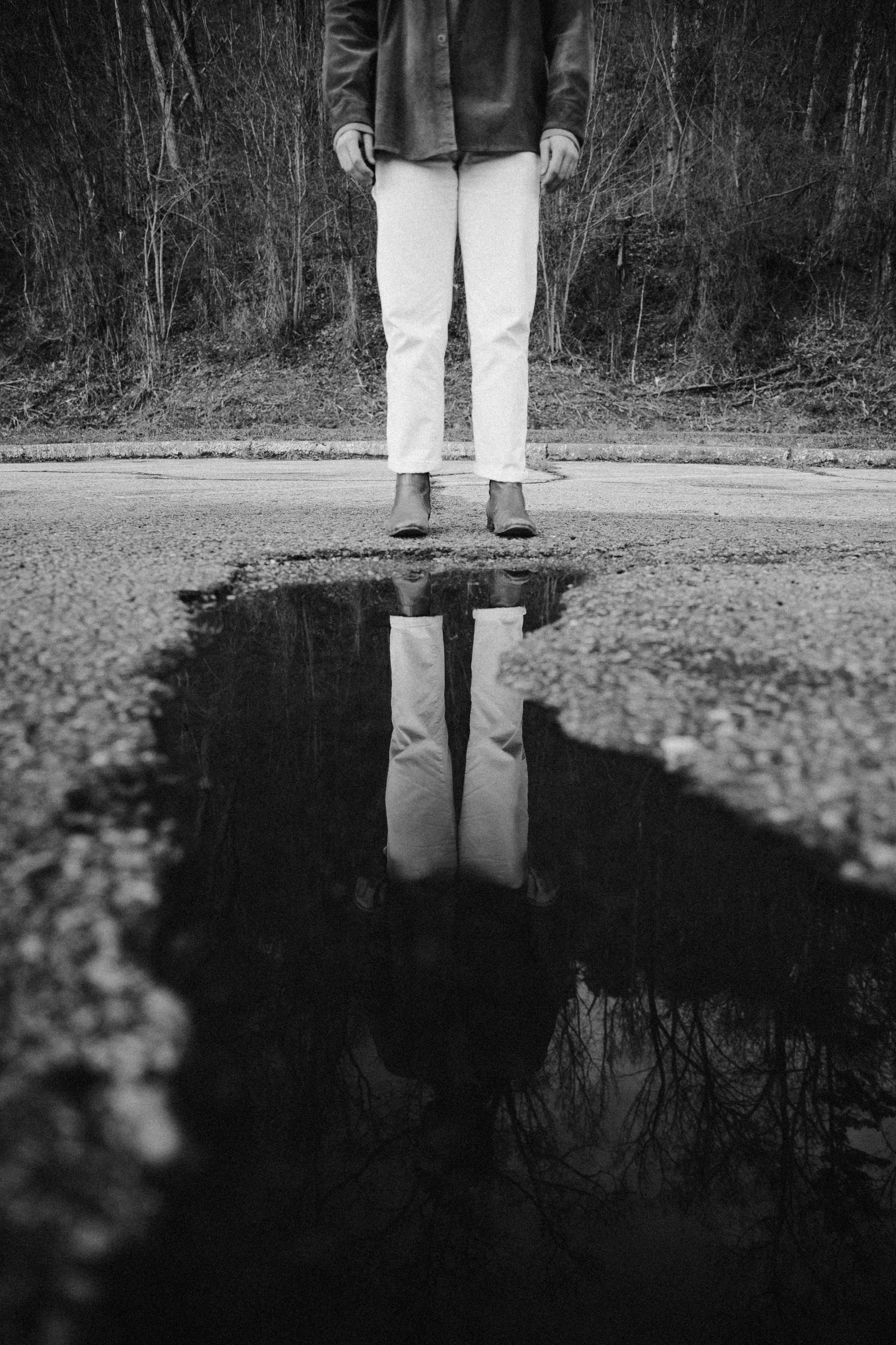 A person stands beside a puddle, their reflection captured in the still water, surrounded by a muted landscape of trees. The scene evokes a sense of introspection.