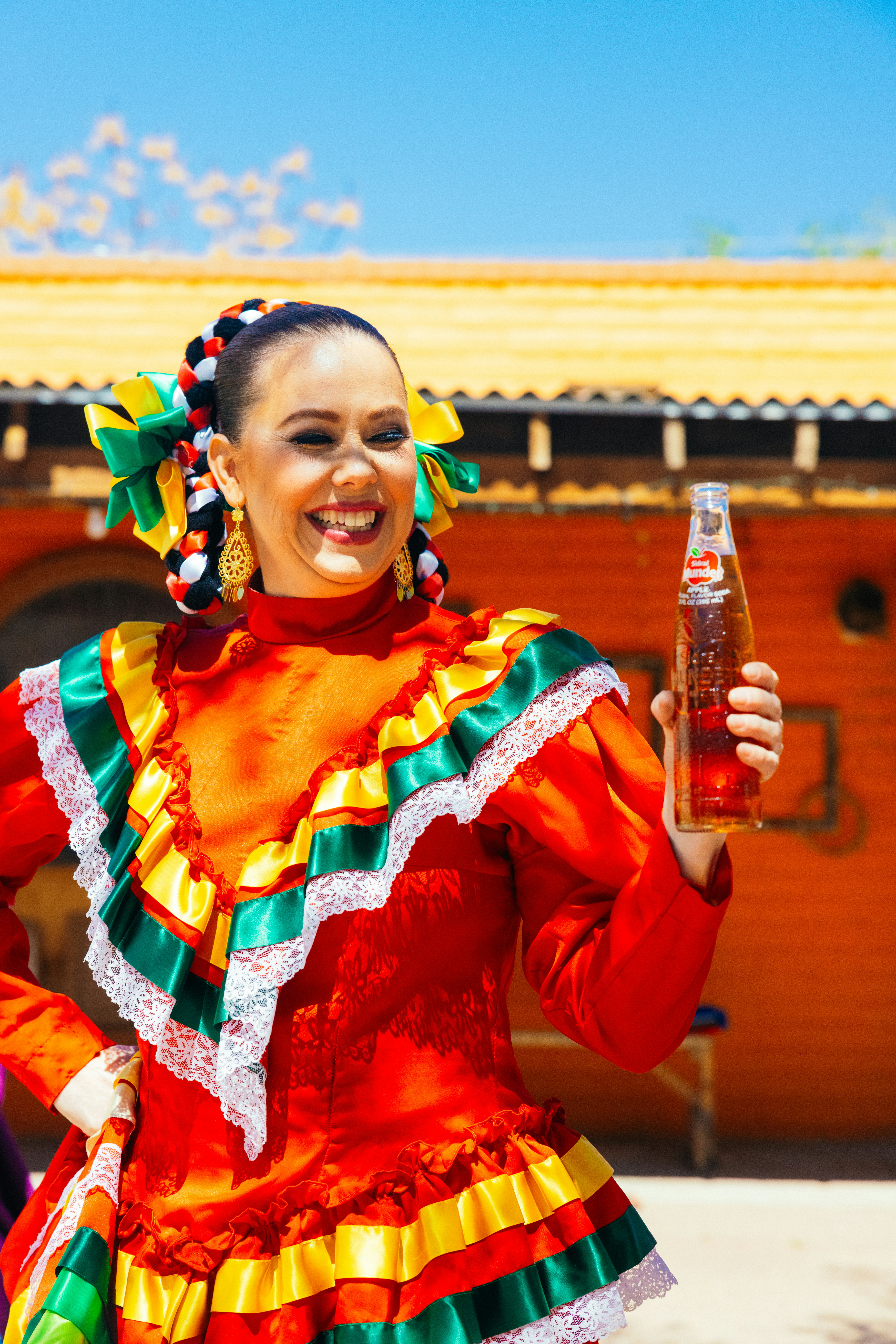 Photograph of a joyful dancer in a bright fiesta dress with multi-colored ribbons, raising a glass bottle outdoors. The background features an orange brick wall.