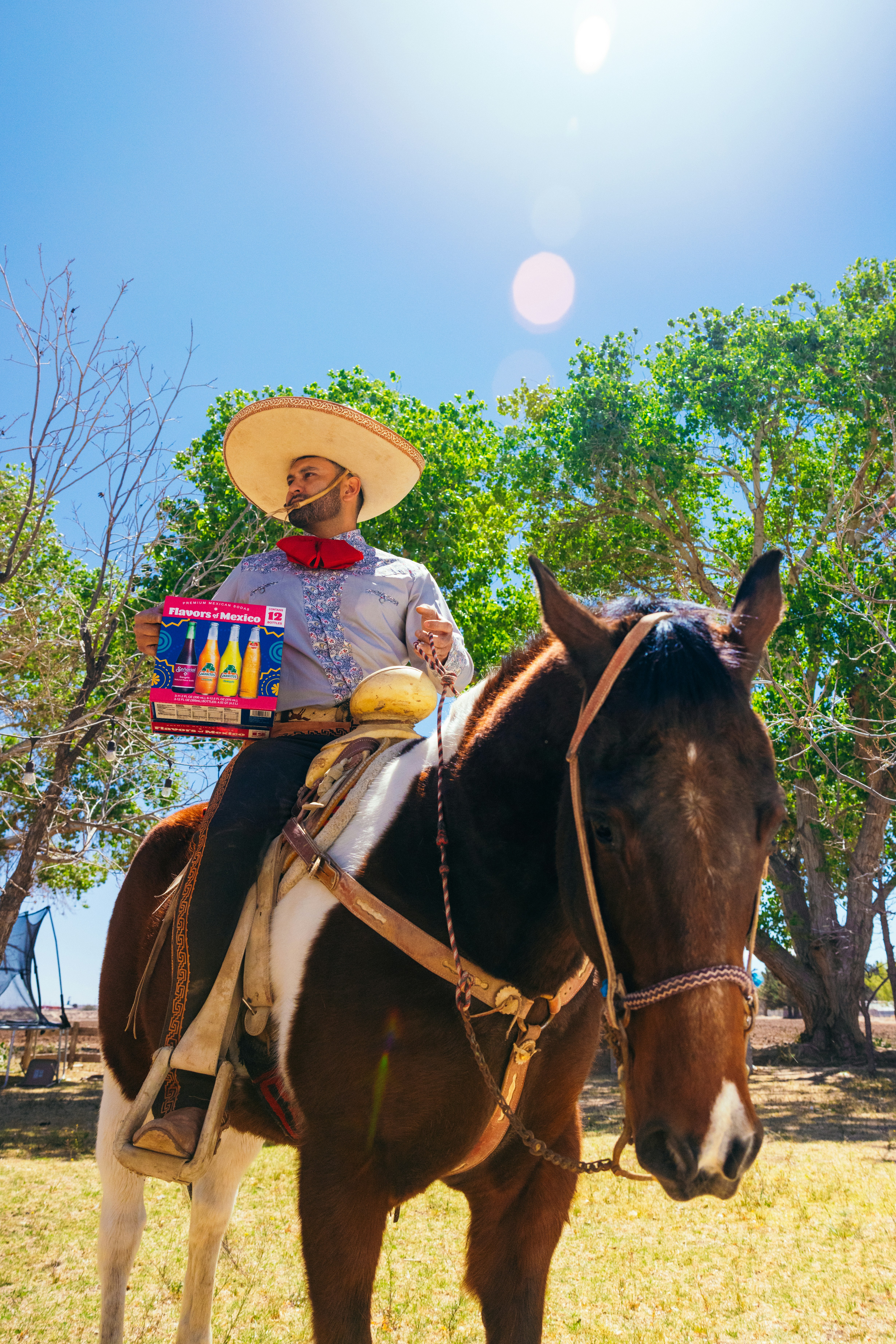 A man in a wide-brim sombrero rides a horse in a sunlit outdoor scene, holding a colorful box of bottles; photograph.