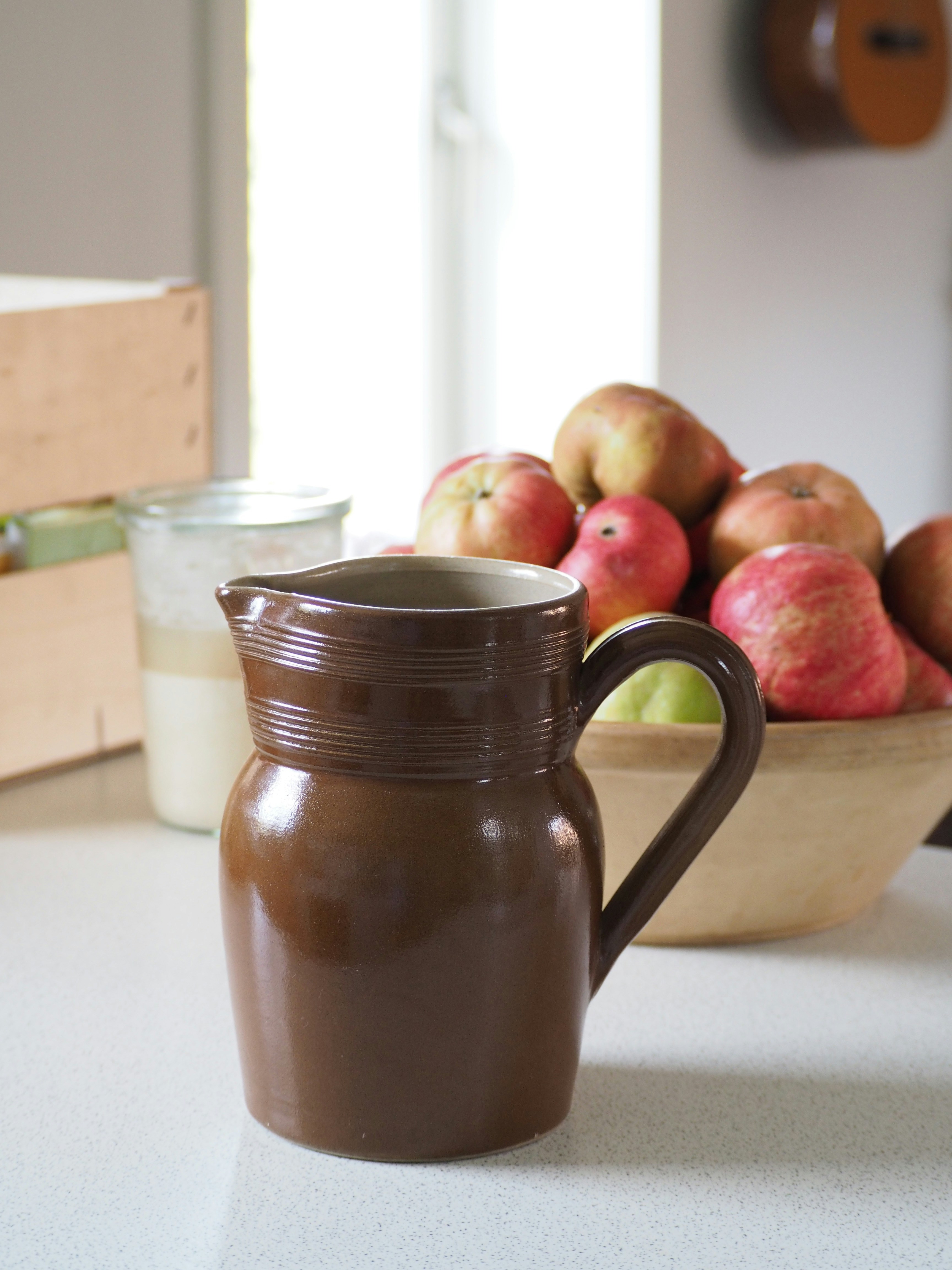 A brown pitcher sitting on top of a counter next to a bowl of apples ...