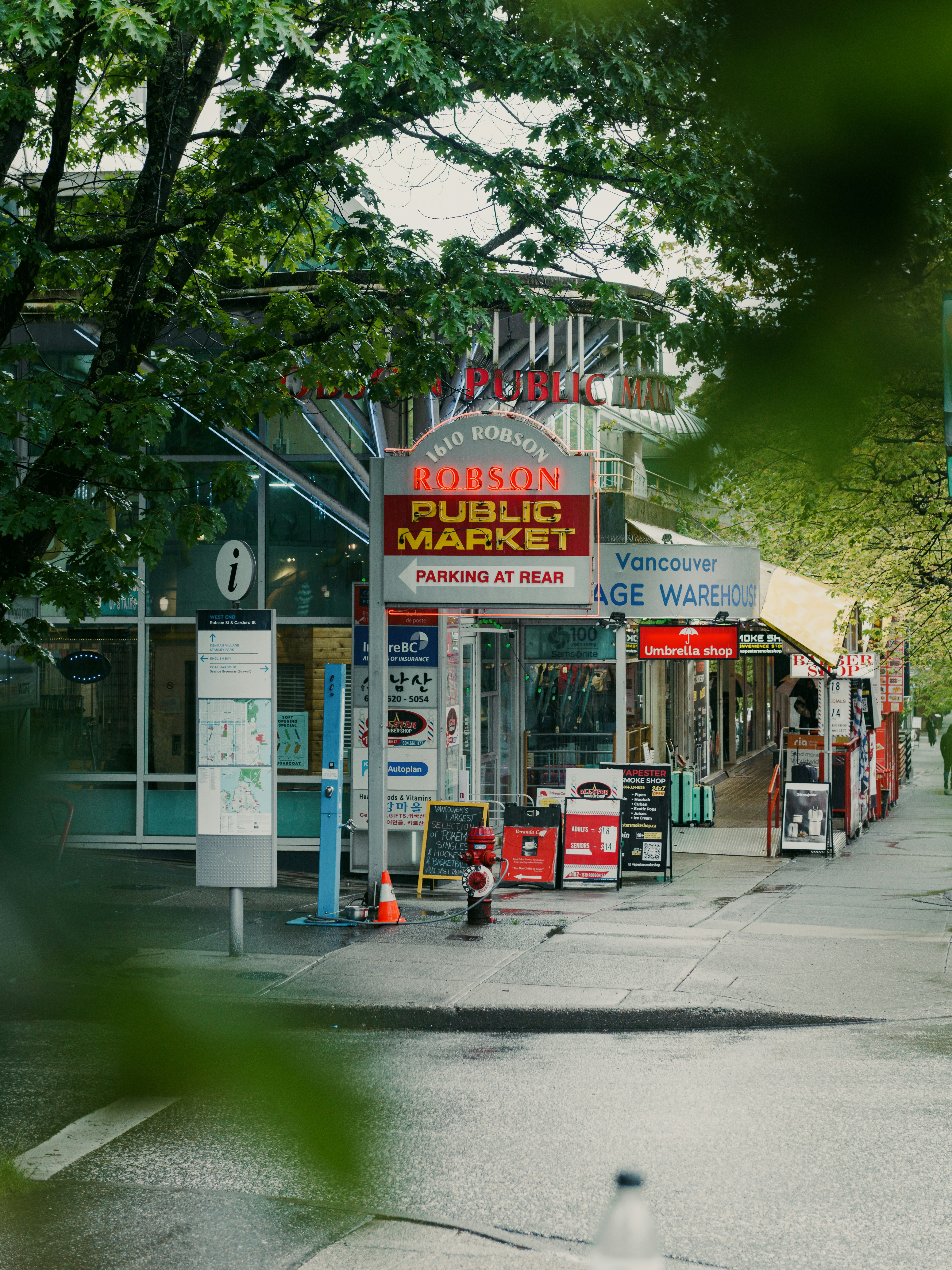 Robson Public Market sign prominently displayed amidst a bustling street scene, framed by lush green foliage. Various storefronts and signage create a vibrant urban atmosphere.