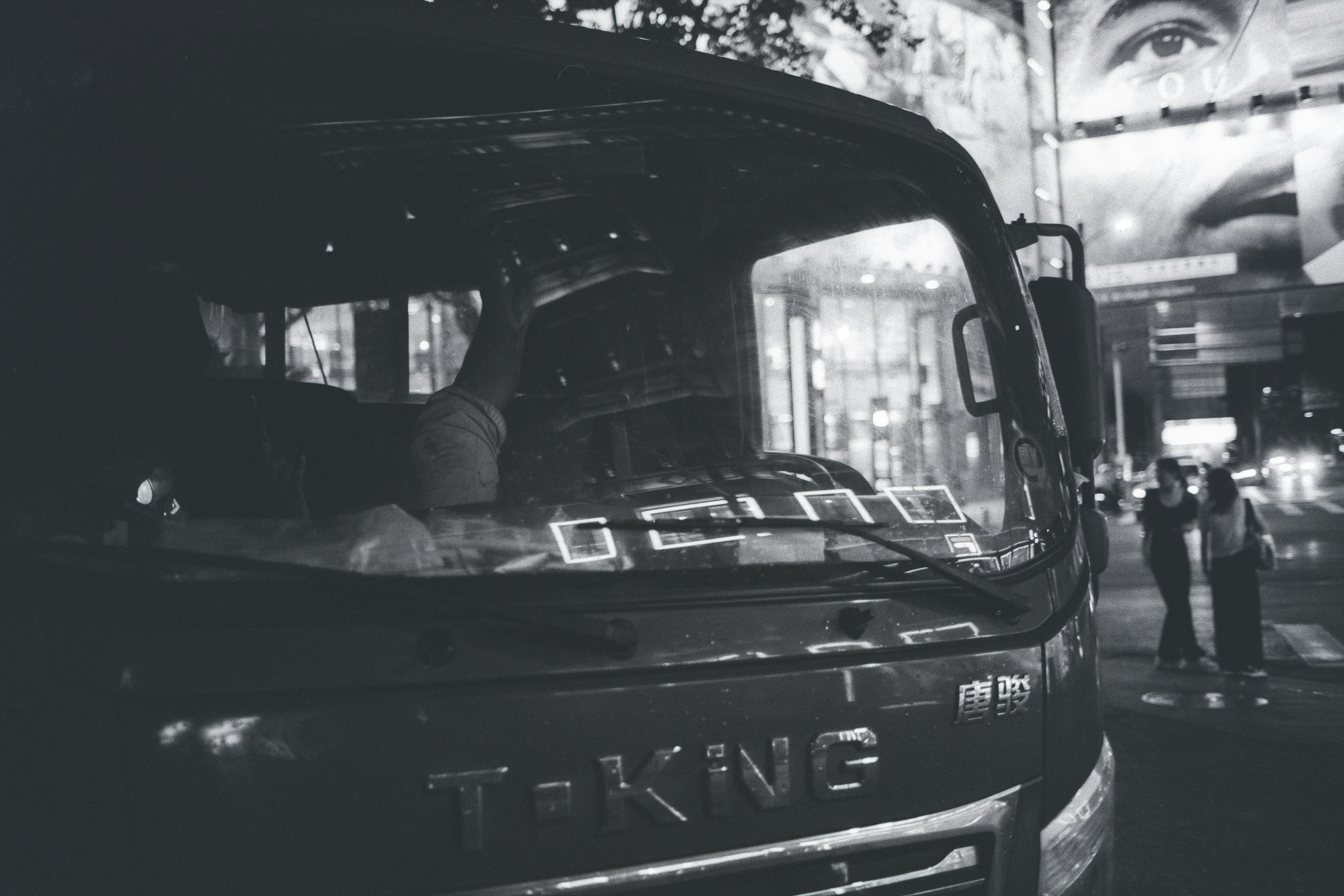 Black-and-white street photograph of a delivery truck cab at night, with glass reflections of city lights in the windshield and pedestrians in the background. The image captures an urban mood rooted in motion and glare on glass.