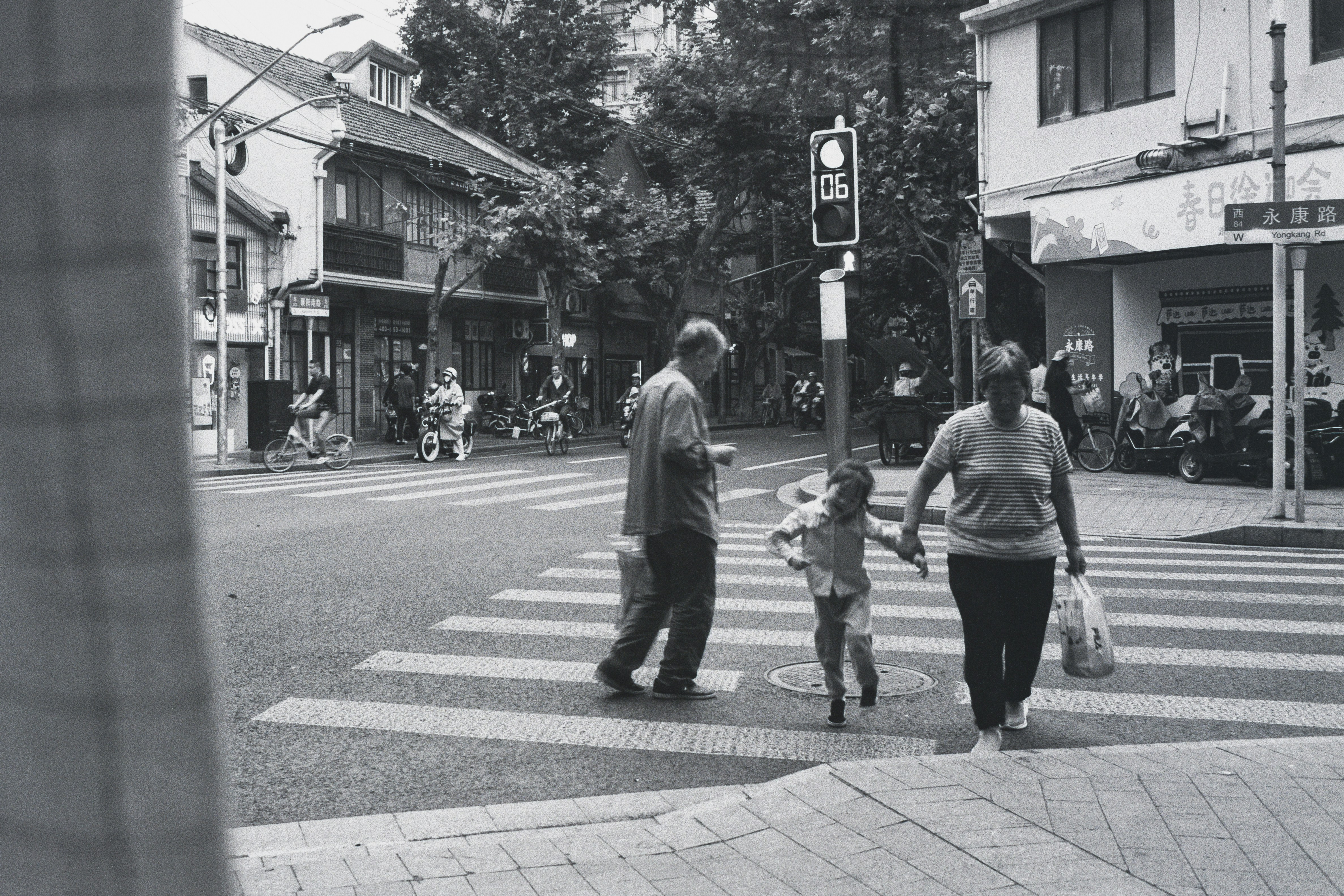 Black-and-white street photograph of a family crossing a busy city intersection at a zebra crosswalk, with scooters and storefronts in the background. The image emphasizes everyday urban motion and human scale.