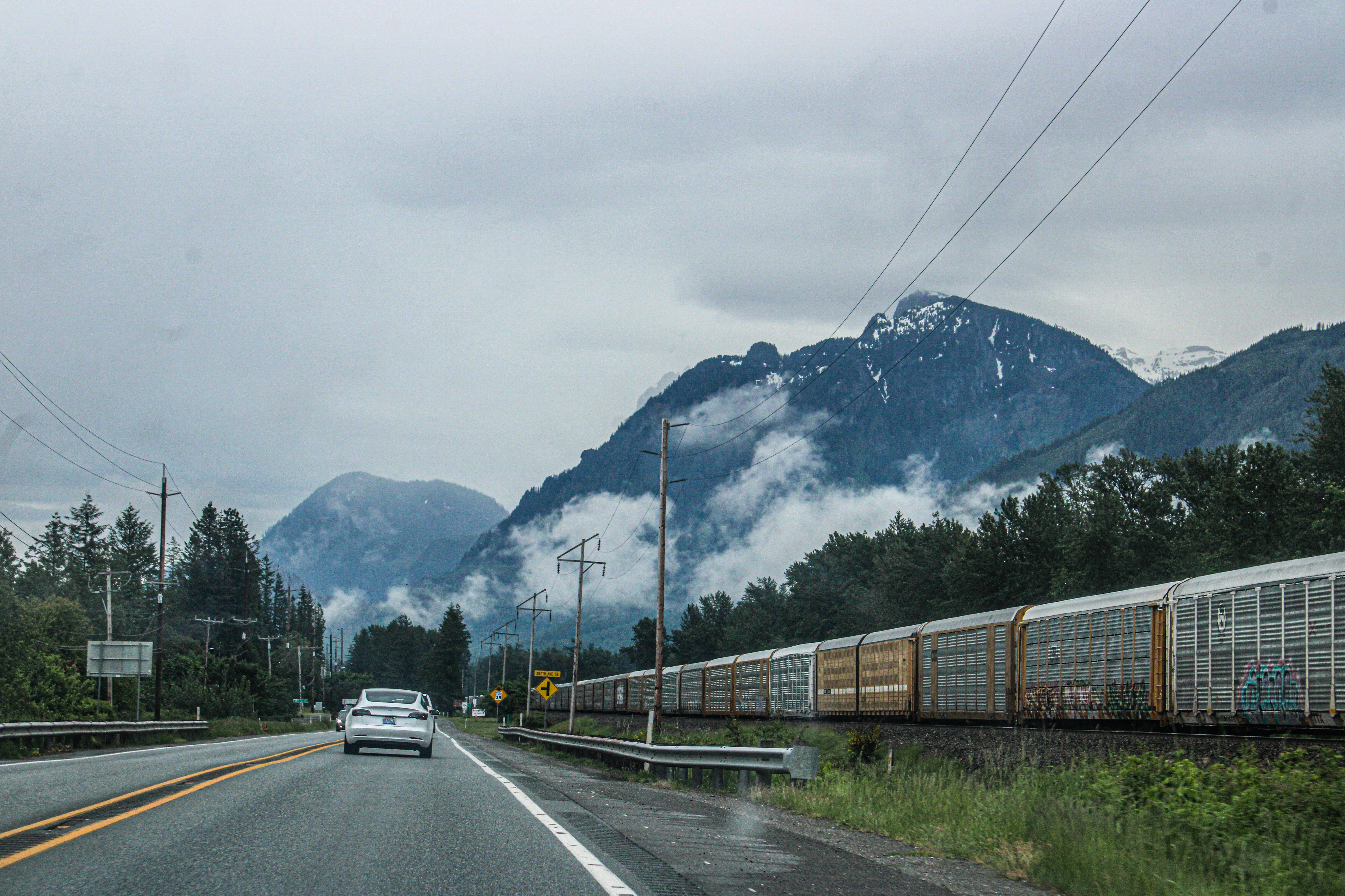 Crossing Lines: A lone white car on a damp road as a mountain-bound freight train threads the valley