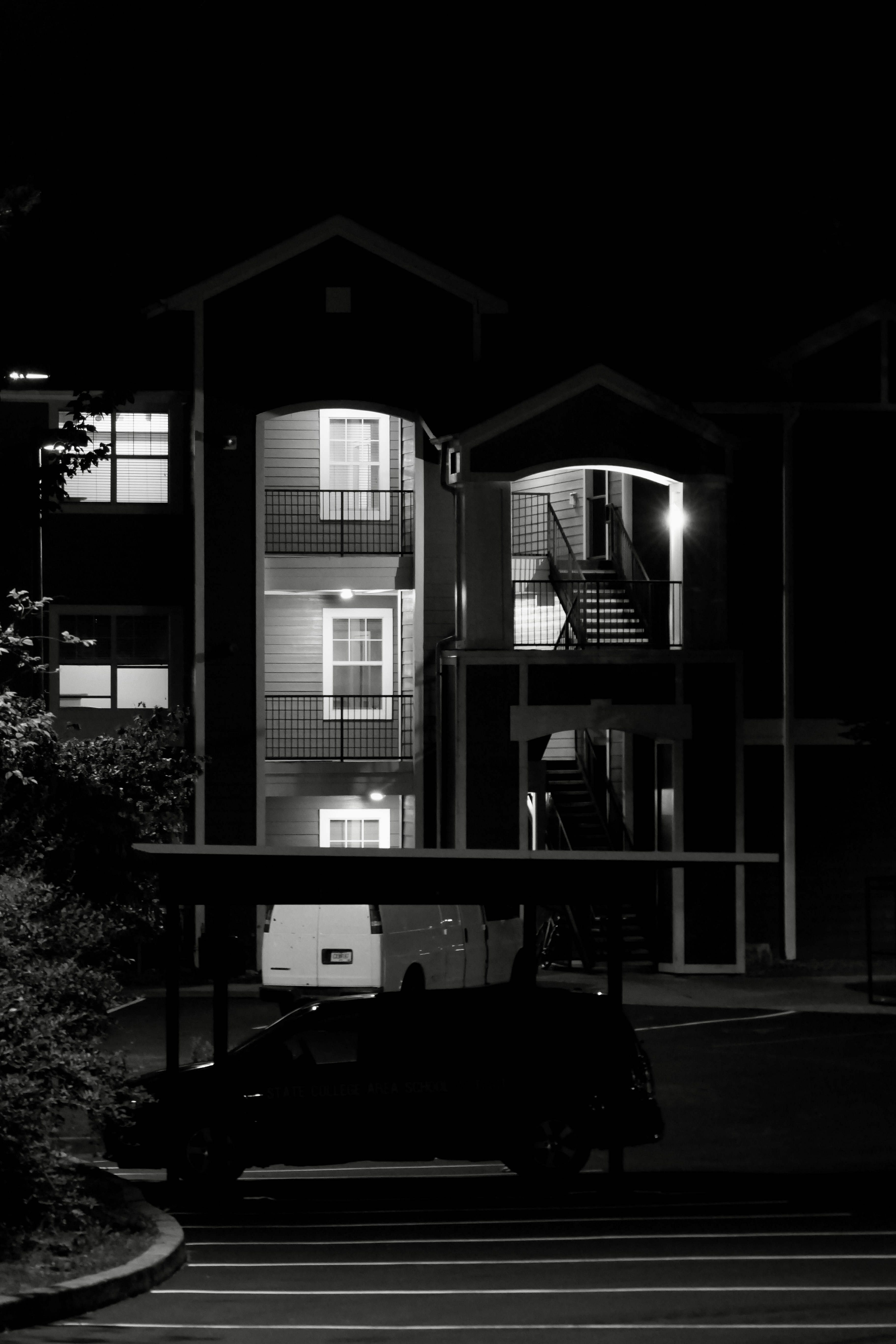 Black-and-white night photograph of a multi-story apartment building with a lit stairwell and a parked van in the foreground.