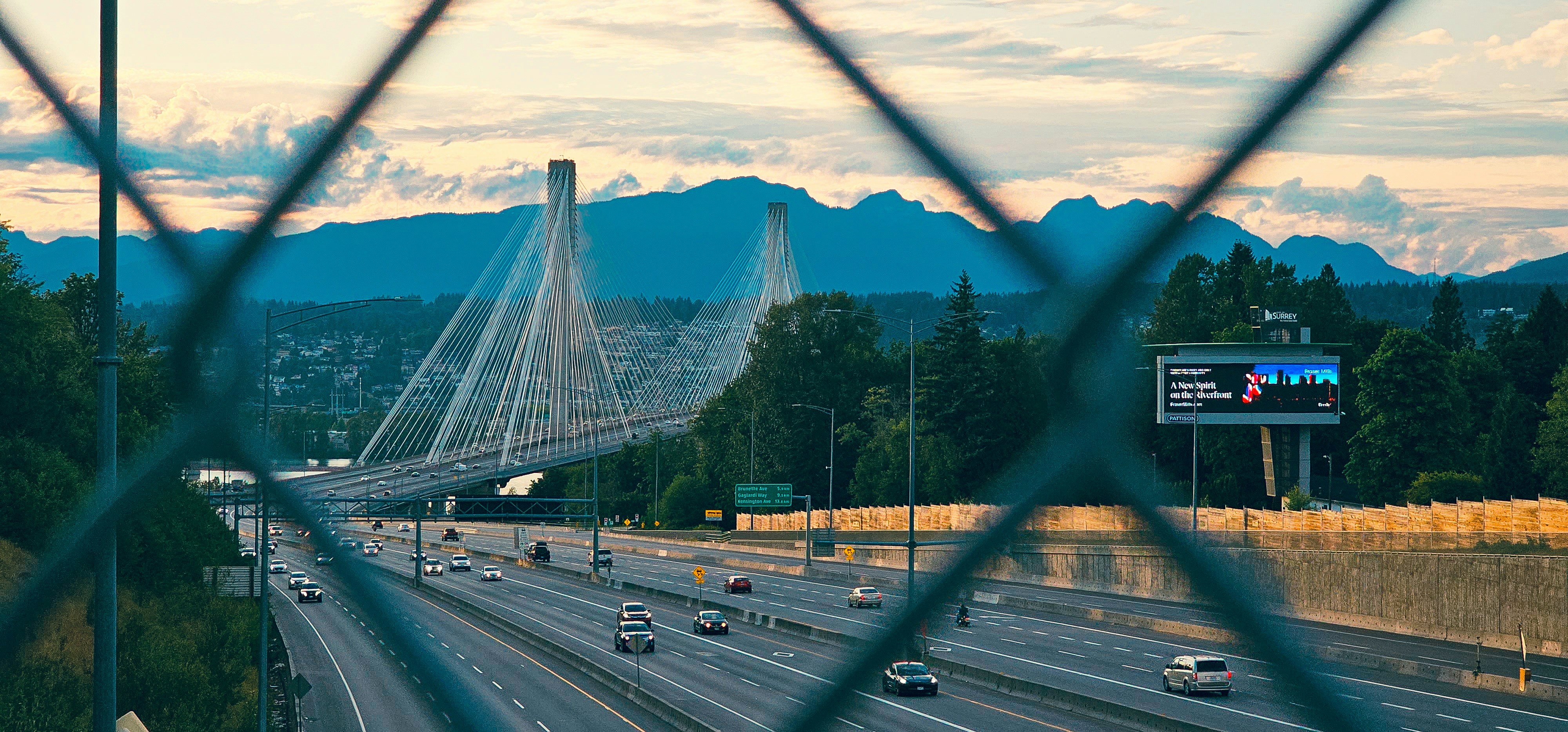 Highway and bridge framed by a chain-link fence with mountains in the background at sunset.