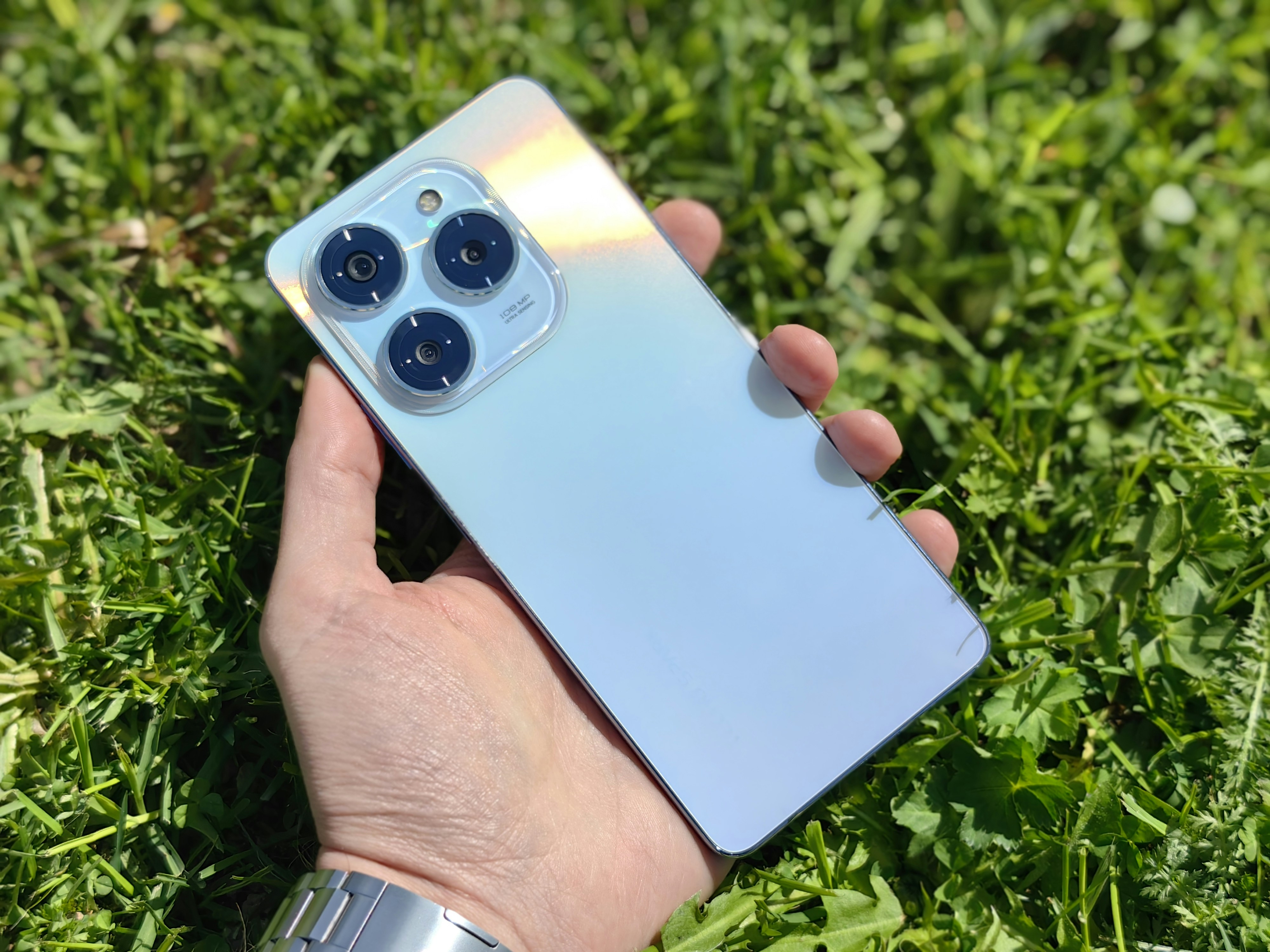 A light-blue smartphone with a prominent triple-camera module rests in a palm over sunlit grass. The reflective back and rounded edges catch the light, highlighting the device’s design.