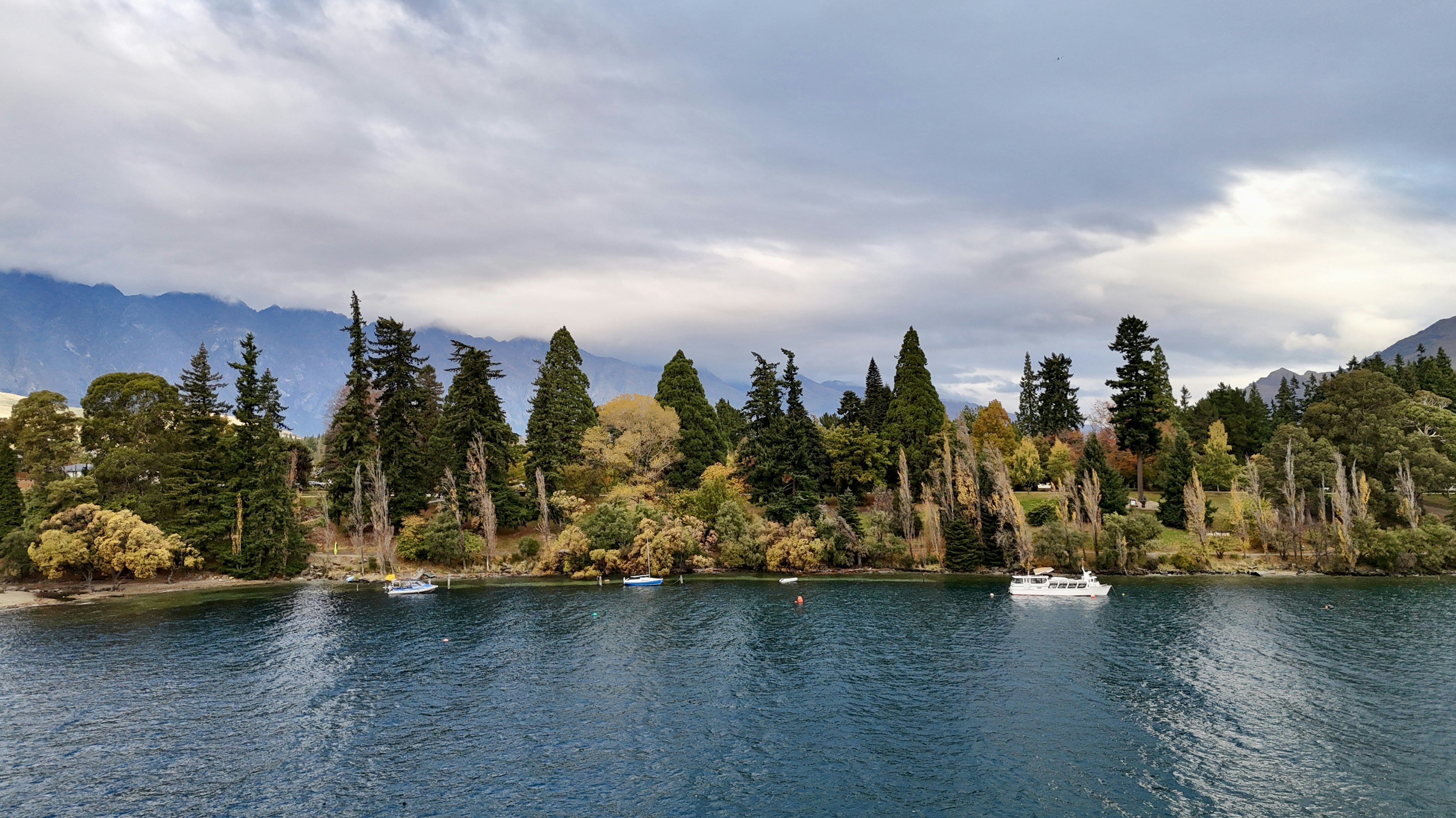 This captivating image captures a tranquil lakeside scene with a rich tapestry of autumn foliage, ranging from vibrant greens to deep oranges and yellows. The calm, deep blue waters reflect the overcast sky, creating a peaceful atmosphere, while the distant mountains add a majestic backdrop. The composition is balanced with the line of trees leading the eye across the frame, making the scene visually striking.