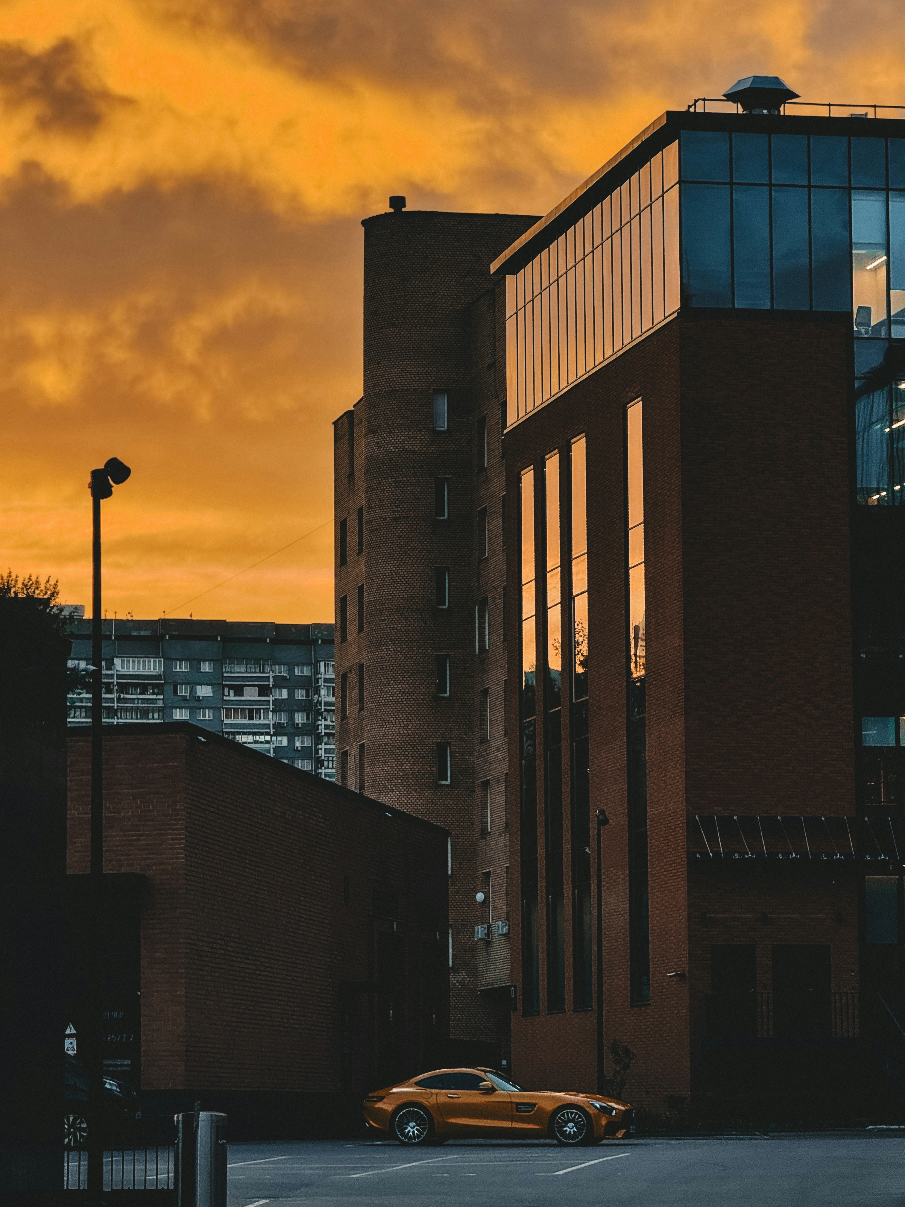 An orange sports car parked in an urban setting, framed by modern brick and glass buildings under a vibrant sunset sky.