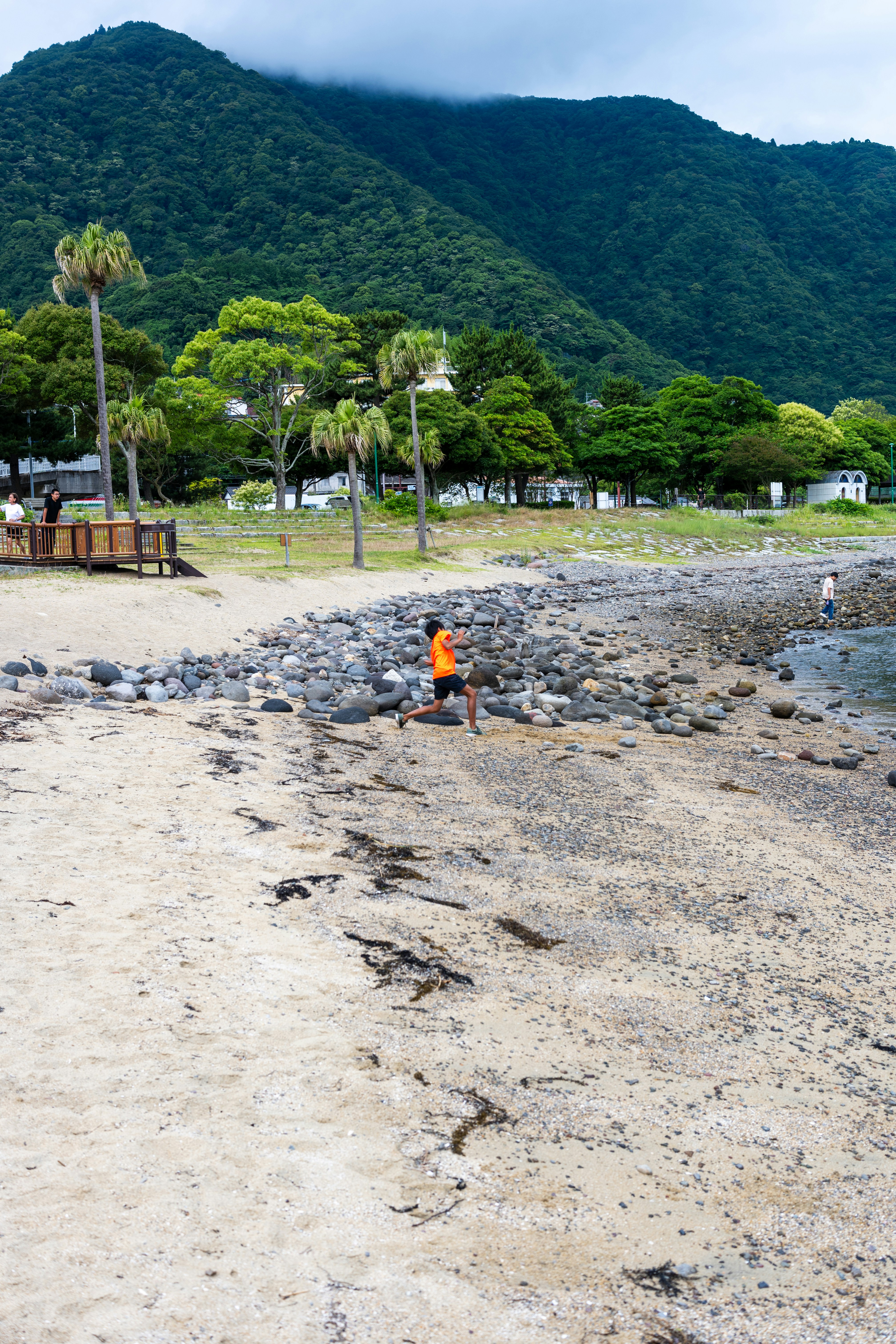 Orange-Clad Figure on a Pebble Beach Beneath Verdant Peaks