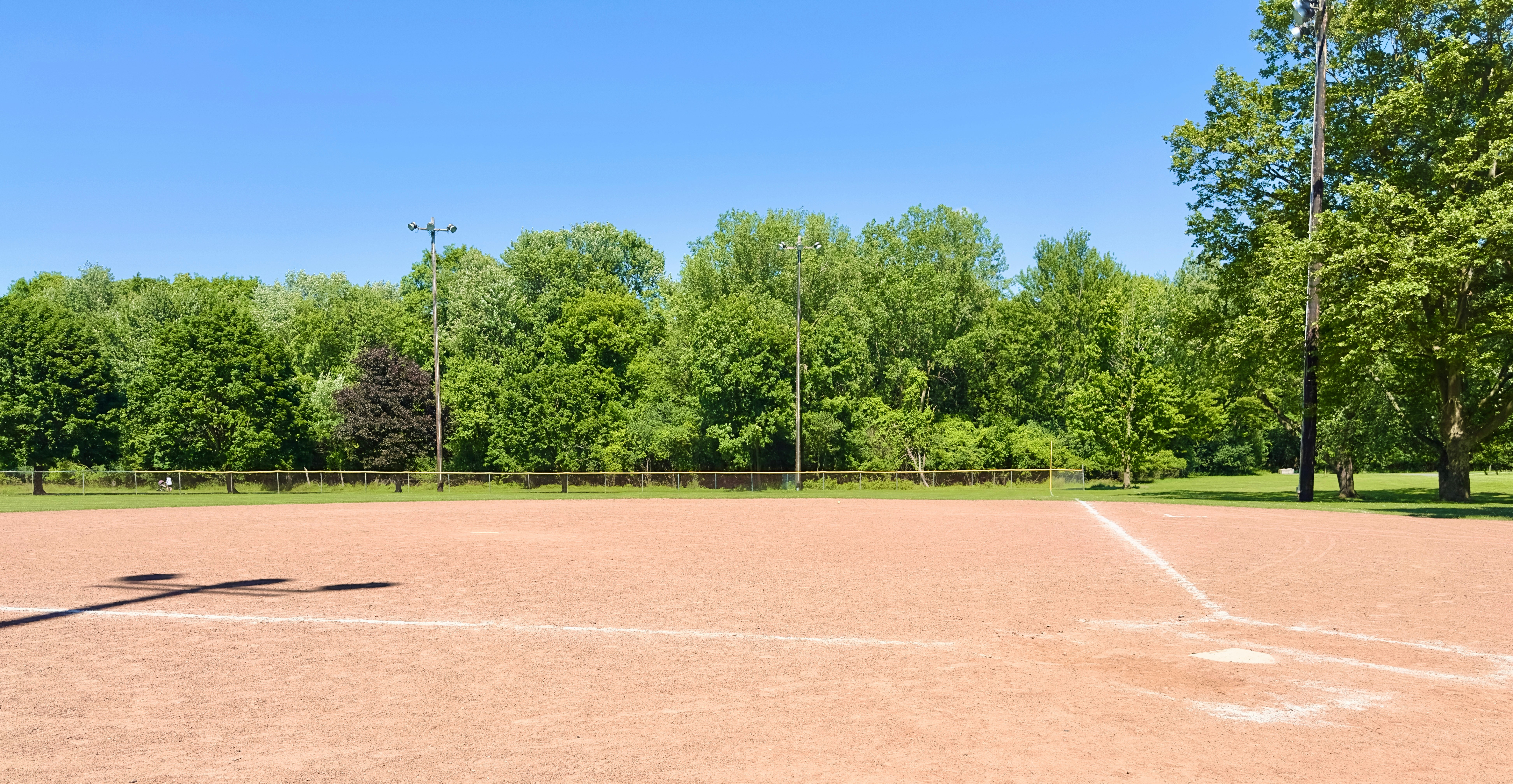 Empty baseball field surrounded by lush green trees under a clear blue sky.