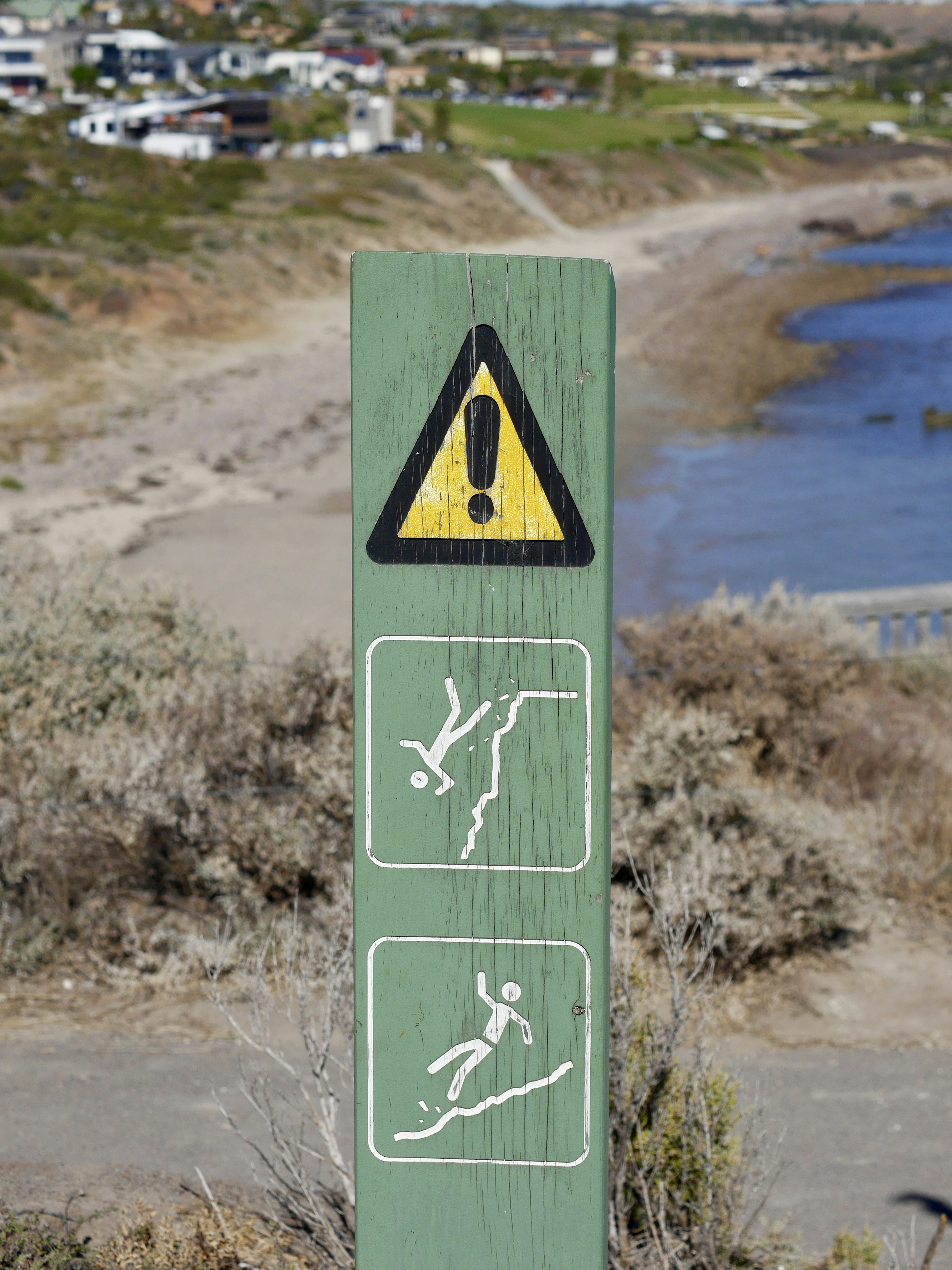 Green wooden post with a triangular caution sign and two hazard pictograms along a coastal trail. The sign indicates falls and slips near the shore.