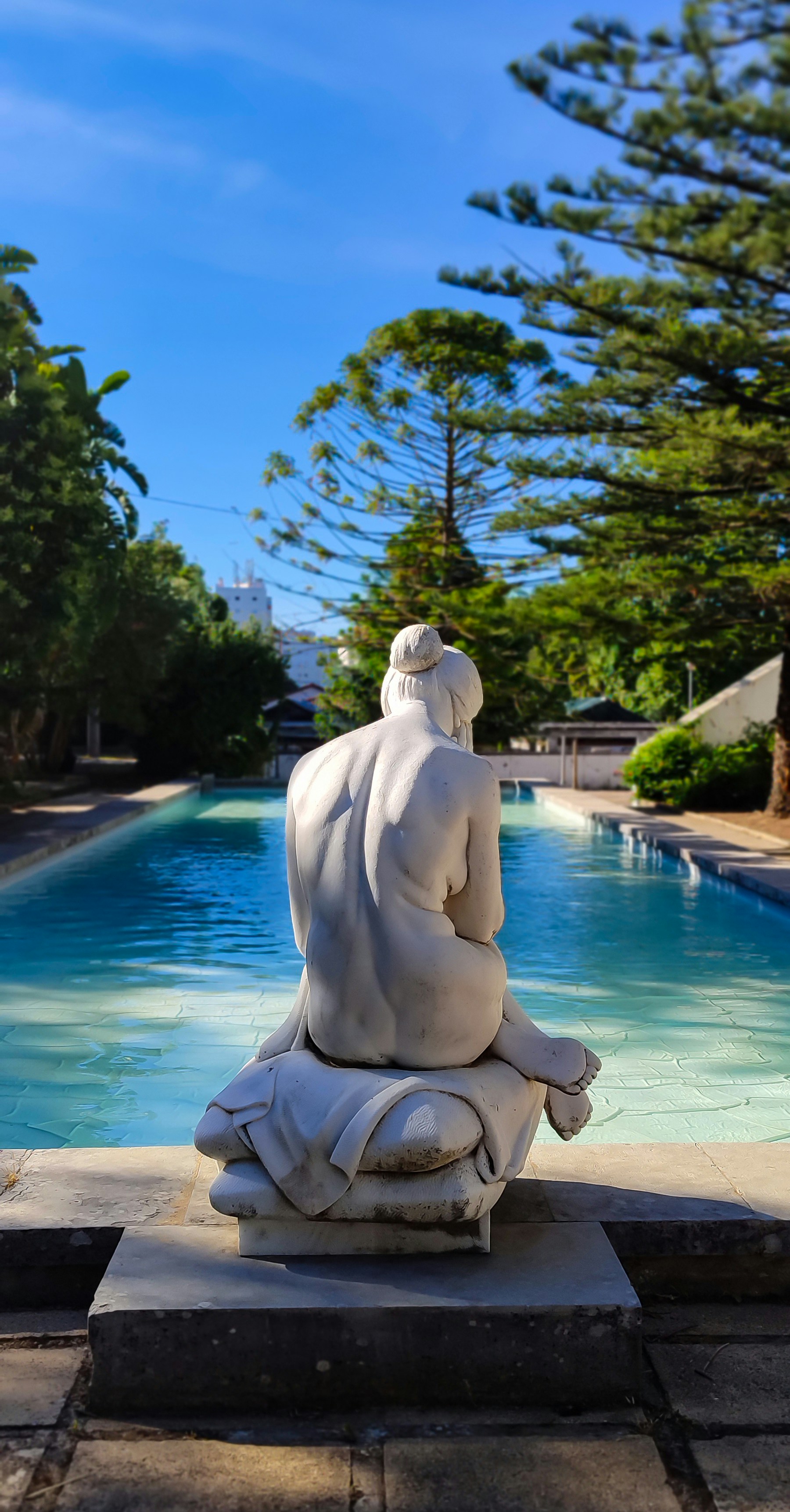 White marble statue seated with its back to the viewer beside a long, sunlit pool. Lush trees frame the scene under a bright blue sky.