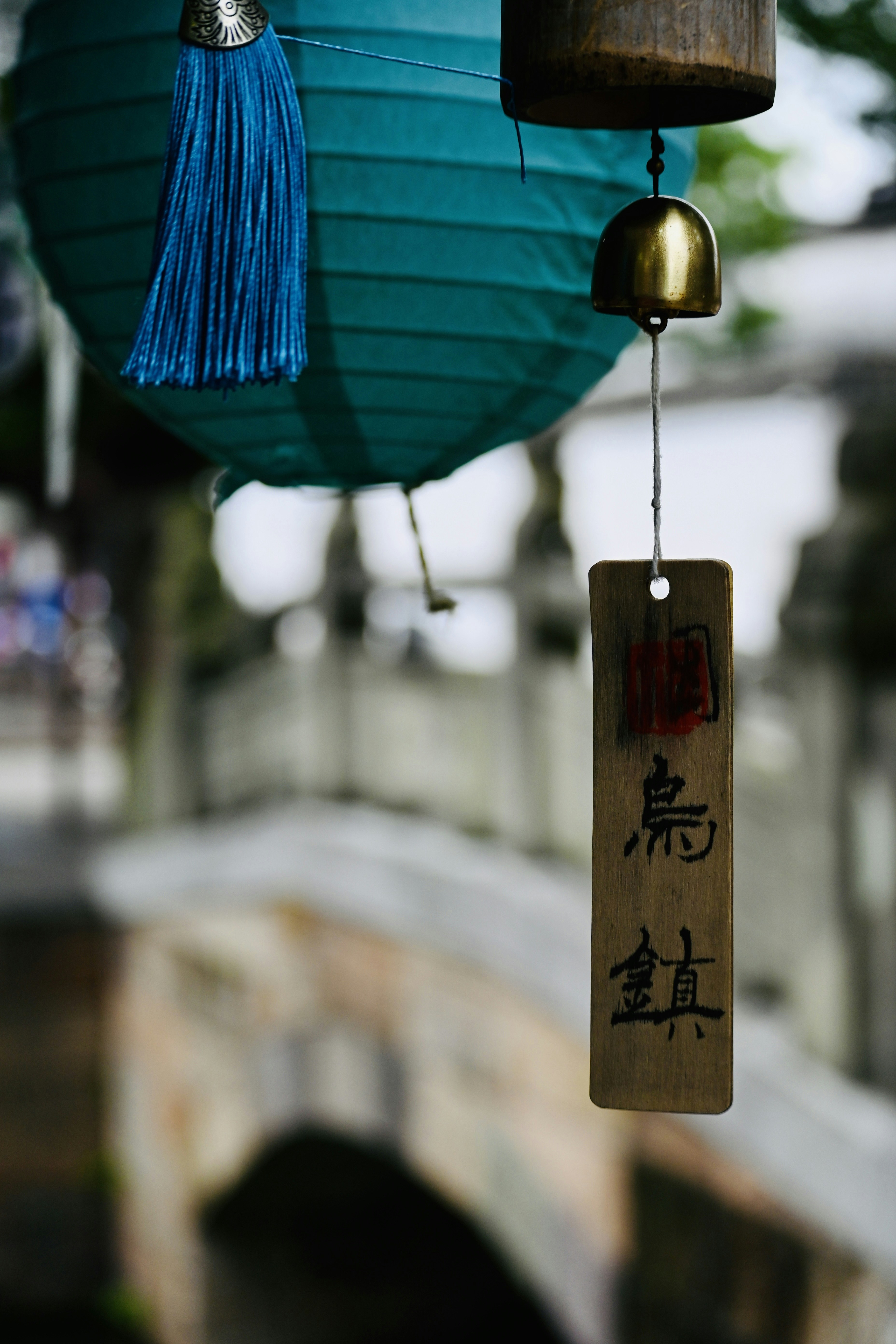 Blue Lantern, Brass Bell, and a Hanging Wooden Talisman on a Quiet Bridge