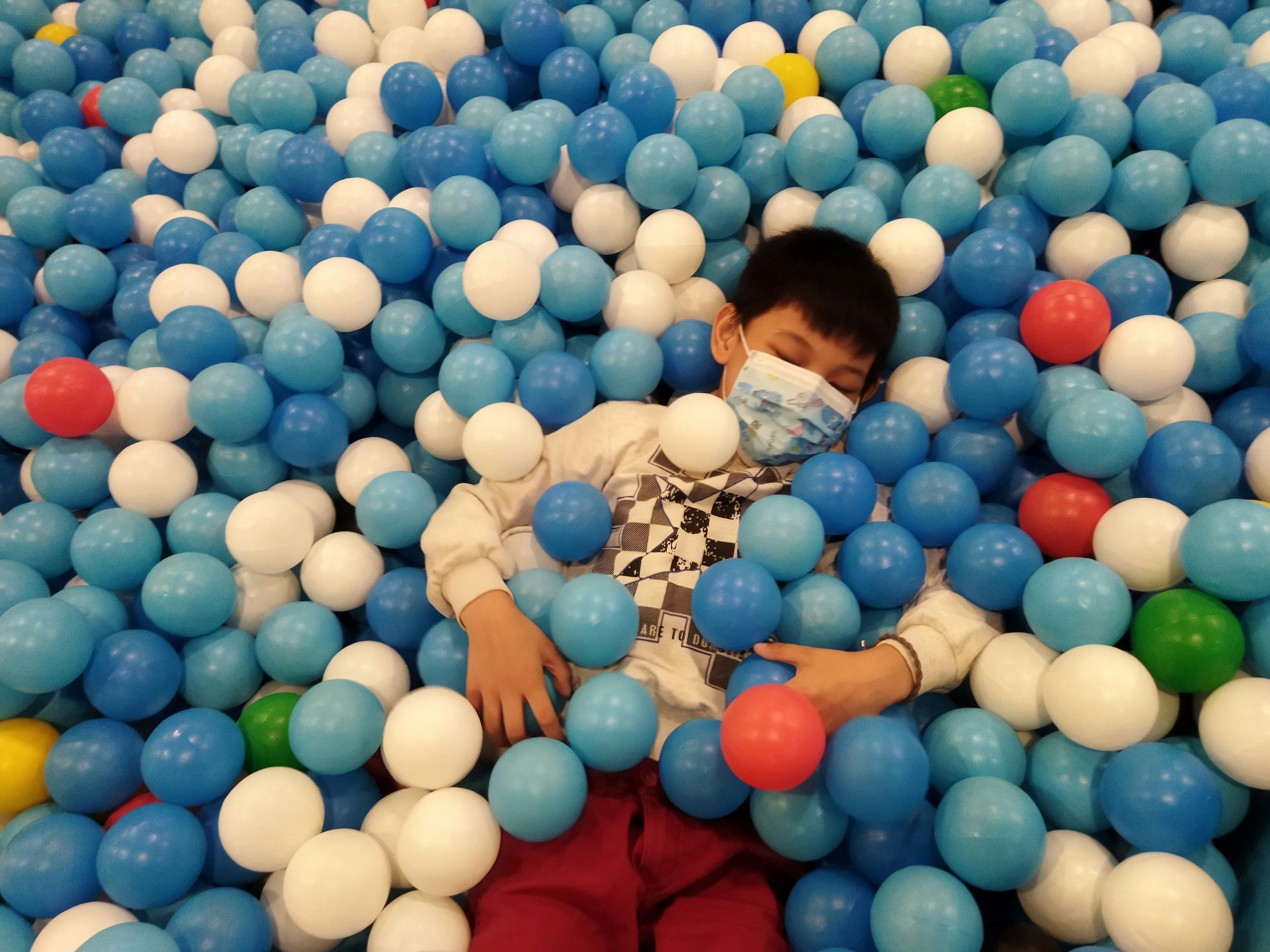 Child relaxed among a vibrant assortment of plastic balls in a play area, wearing a mask. The scene captures a moment of playful tranquility.