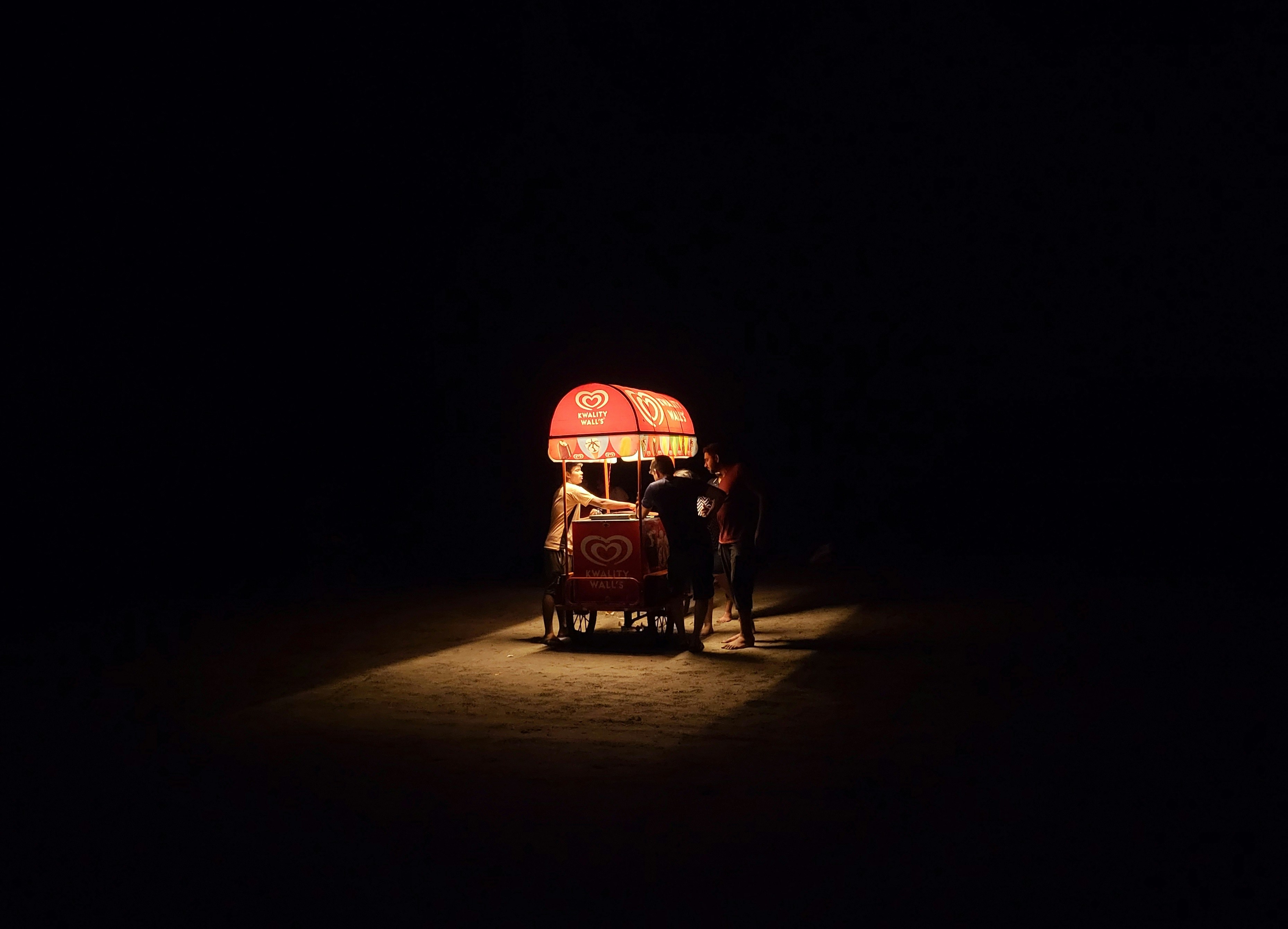 Night-time photograph of a small vendor cart under a red umbrella, illuminated by a warm beam on a dark beach.