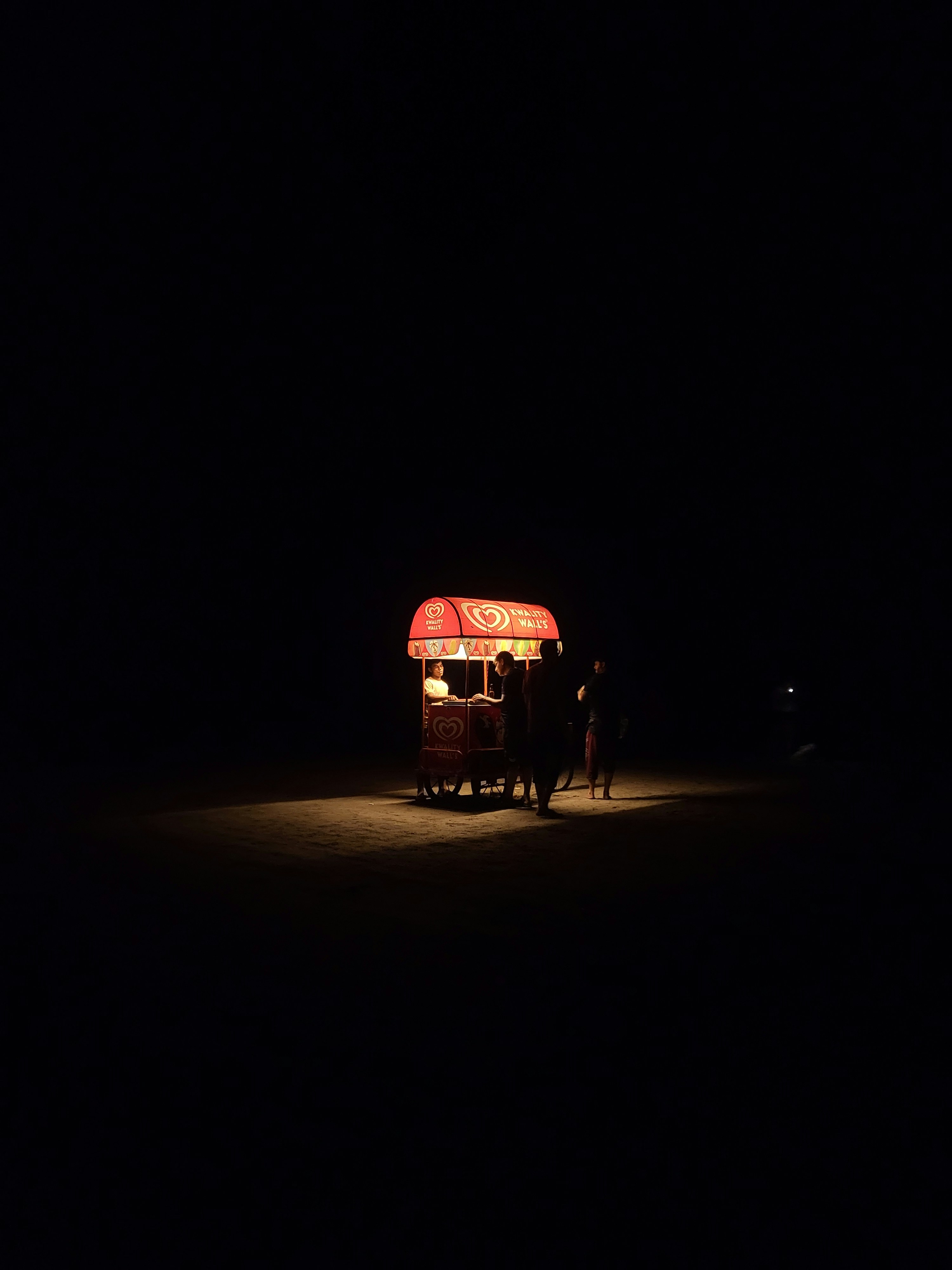 Photograph of a small red-lit cart glowing in the dark, with a few silhouettes gathered around on a quiet beach.