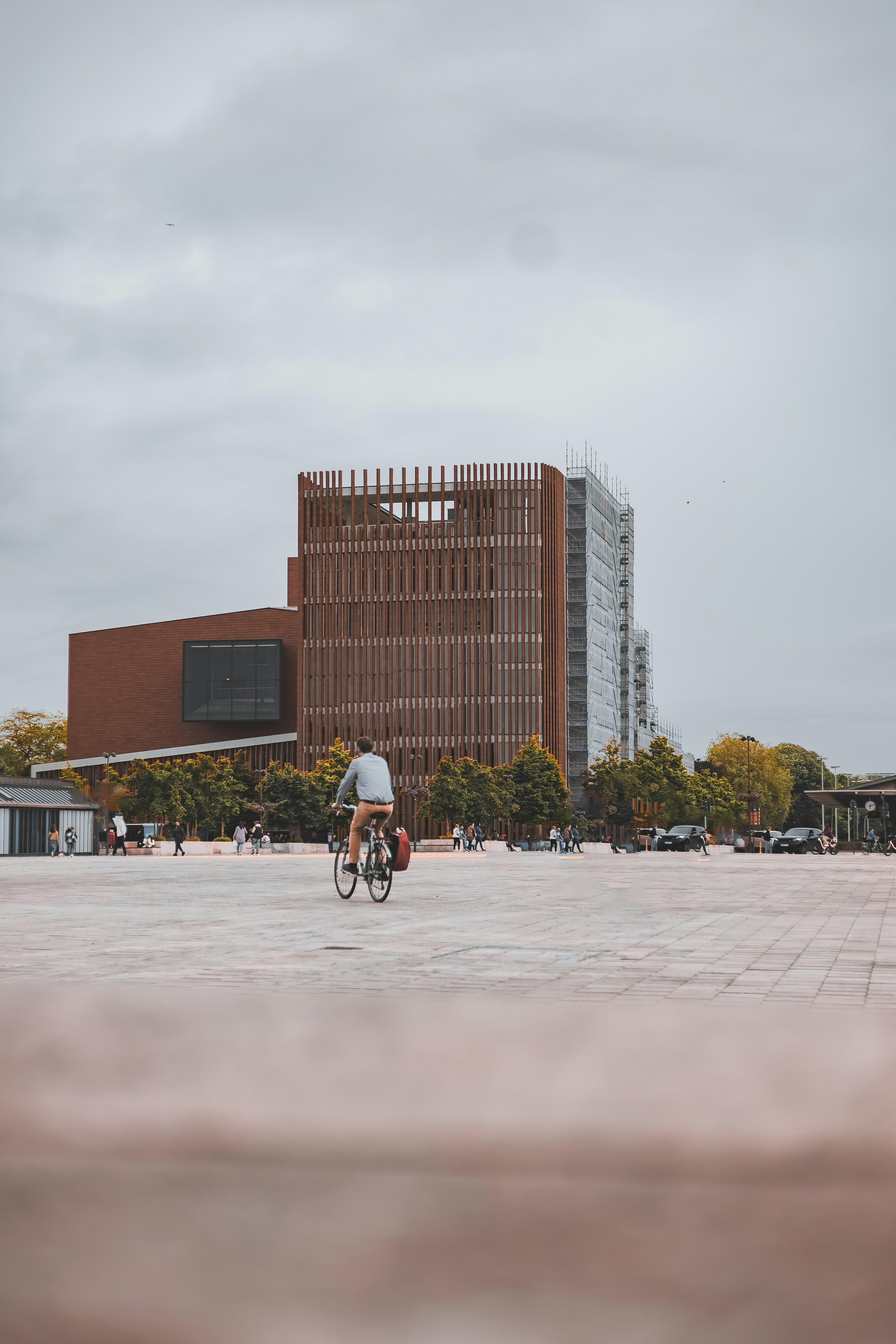 A lone cyclist rides across a wide plaza toward a modern red-brick building, with pedestrians and trees in the background. This is a candid street scene captured in an urban square.