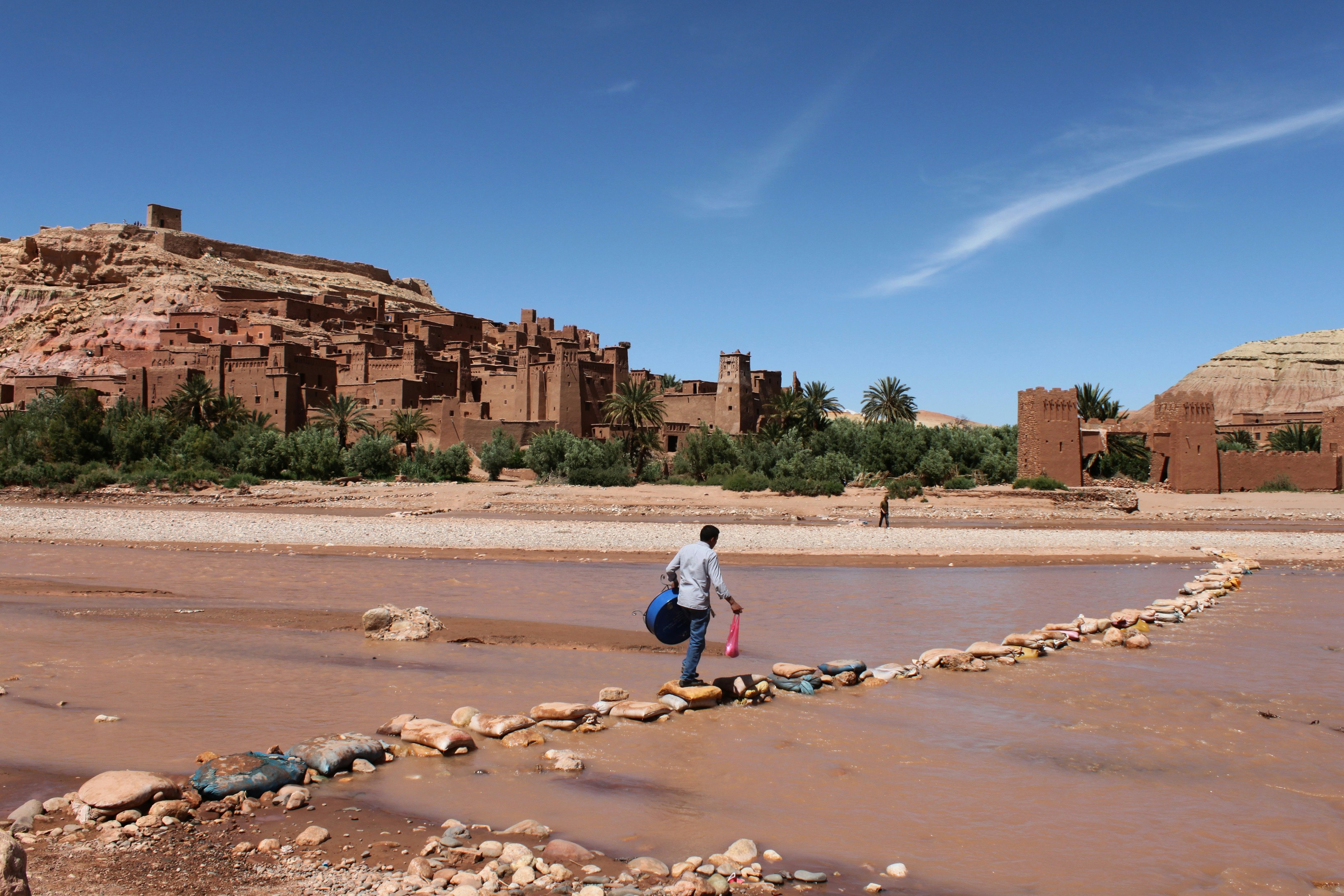 Person crossing a stone pathway over a shallow river with historic adobe structures in the background under a clear blue sky.