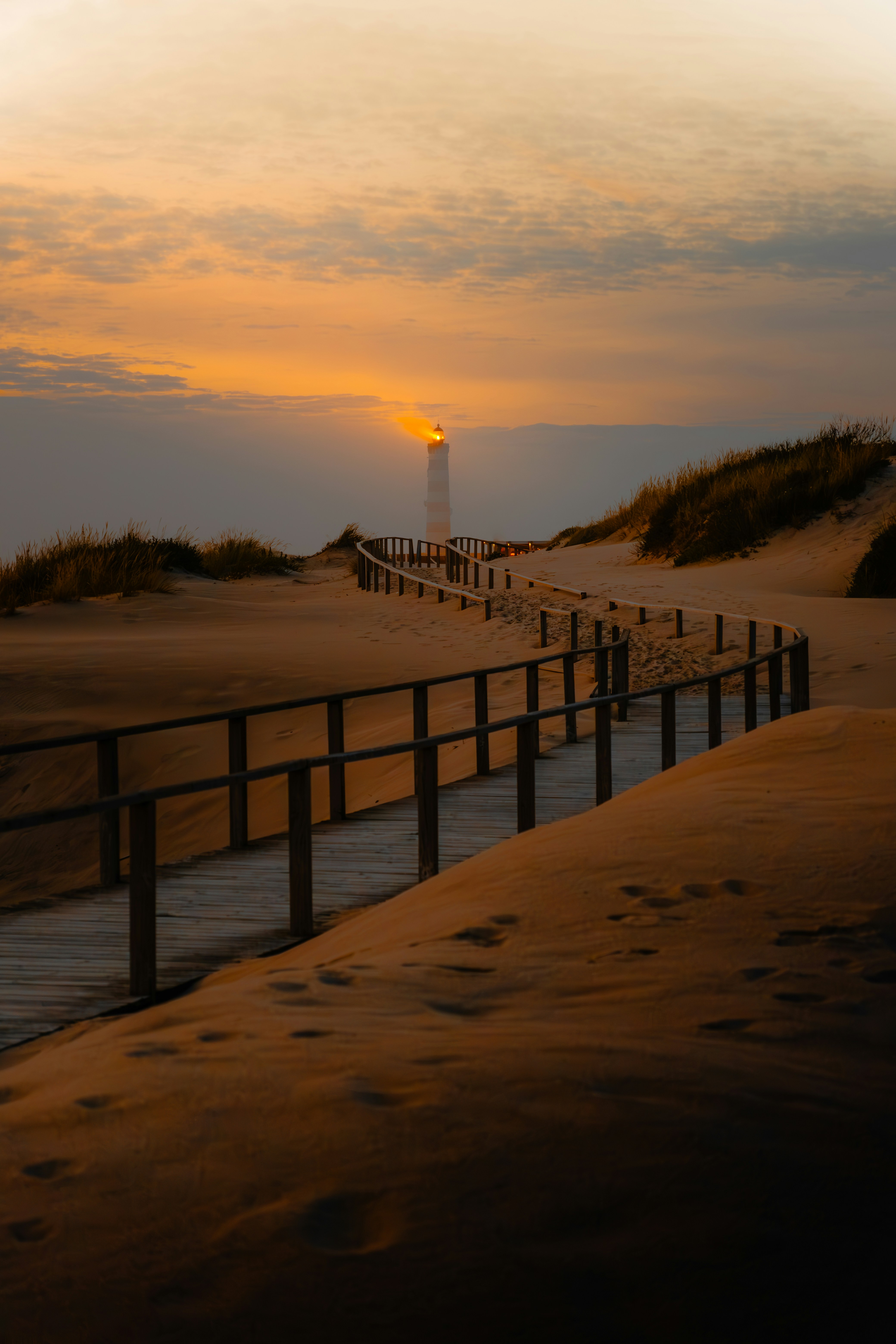 Lighthouse illuminating a sandy boardwalk at sunset with dramatic skies.