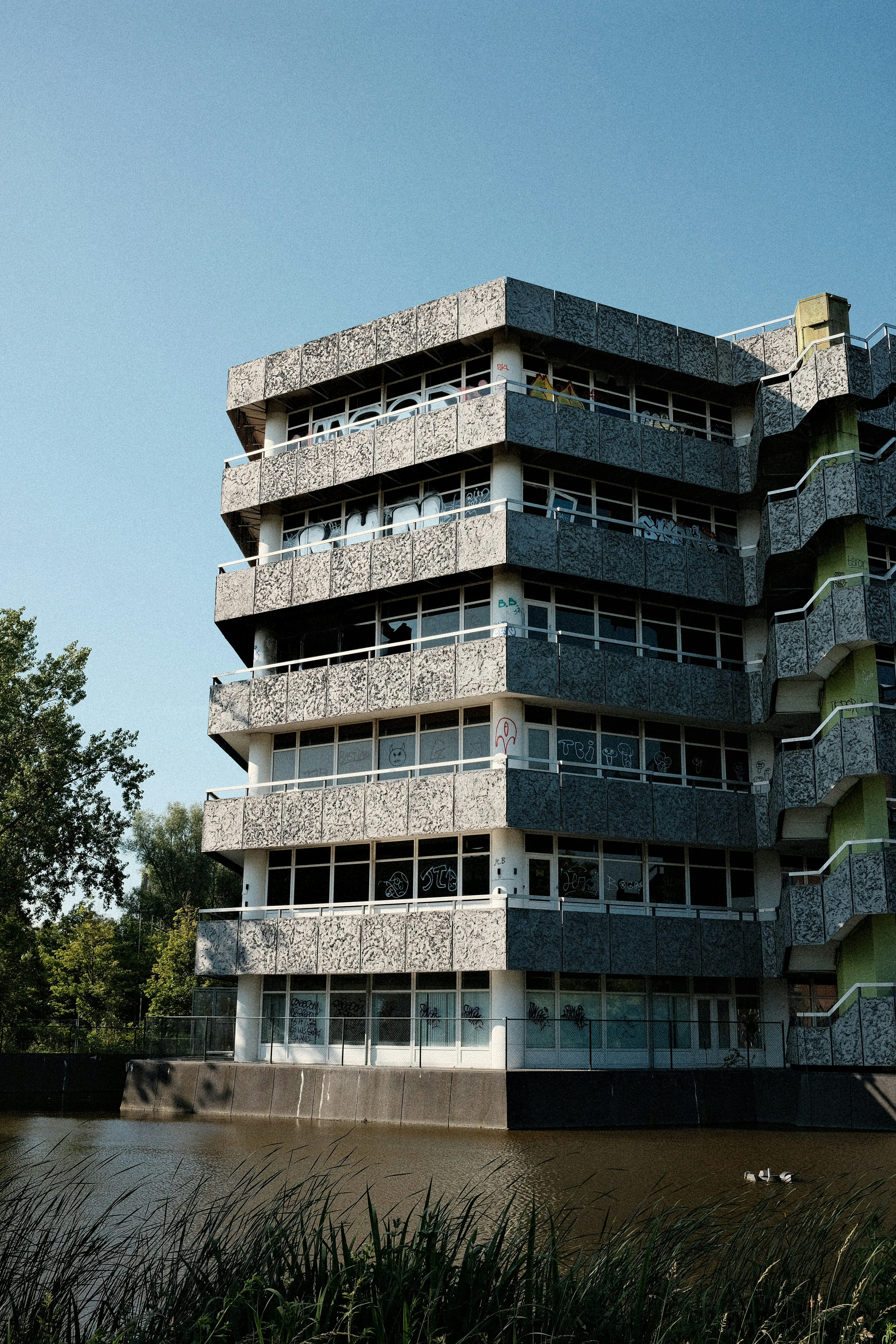 Brutalist building with stacked balconies sits beside a calm canal, under a clear blue sky.