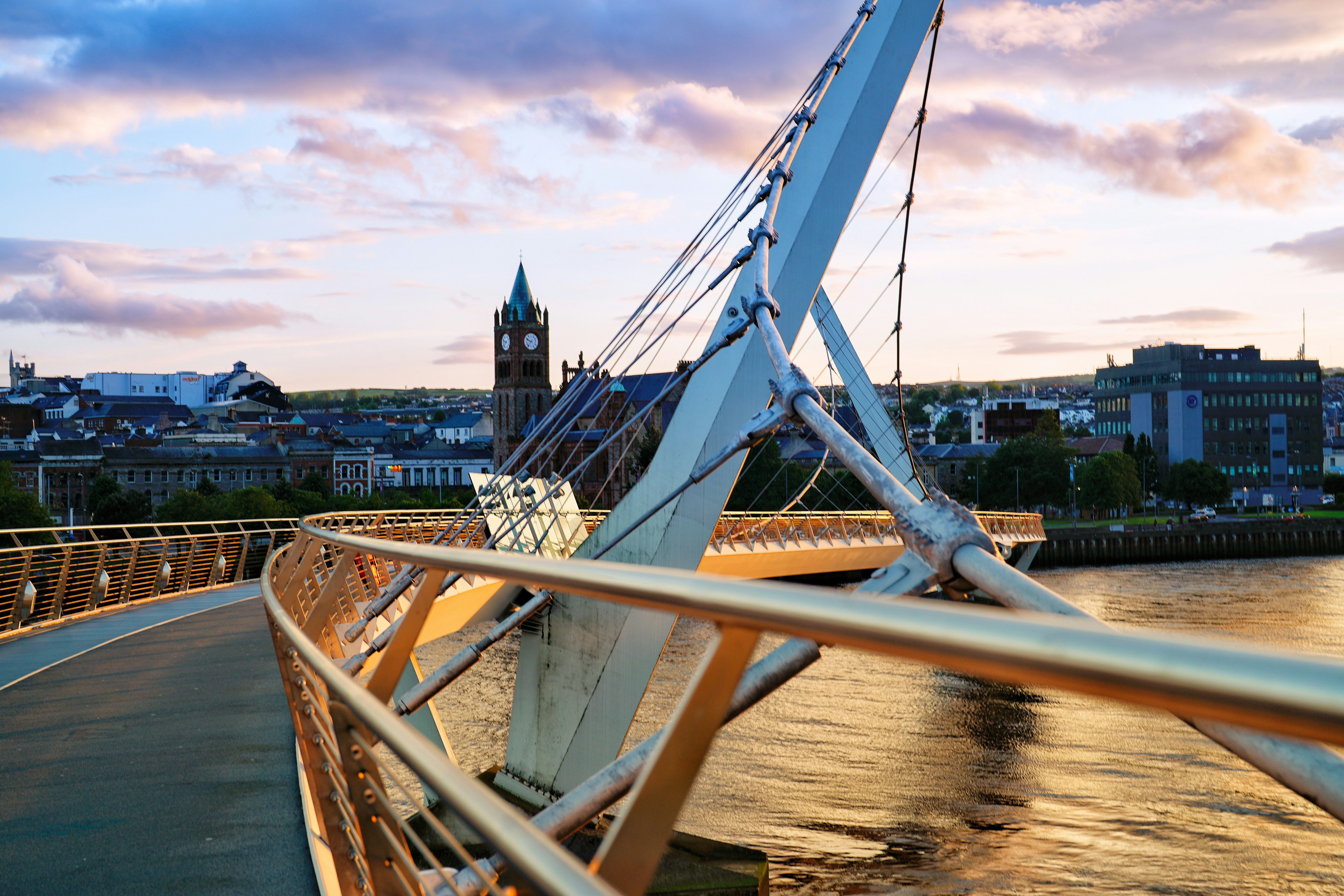 Modern suspension bridge arches gracefully over a river against a vibrant sunset sky, with a cityscape in the background.