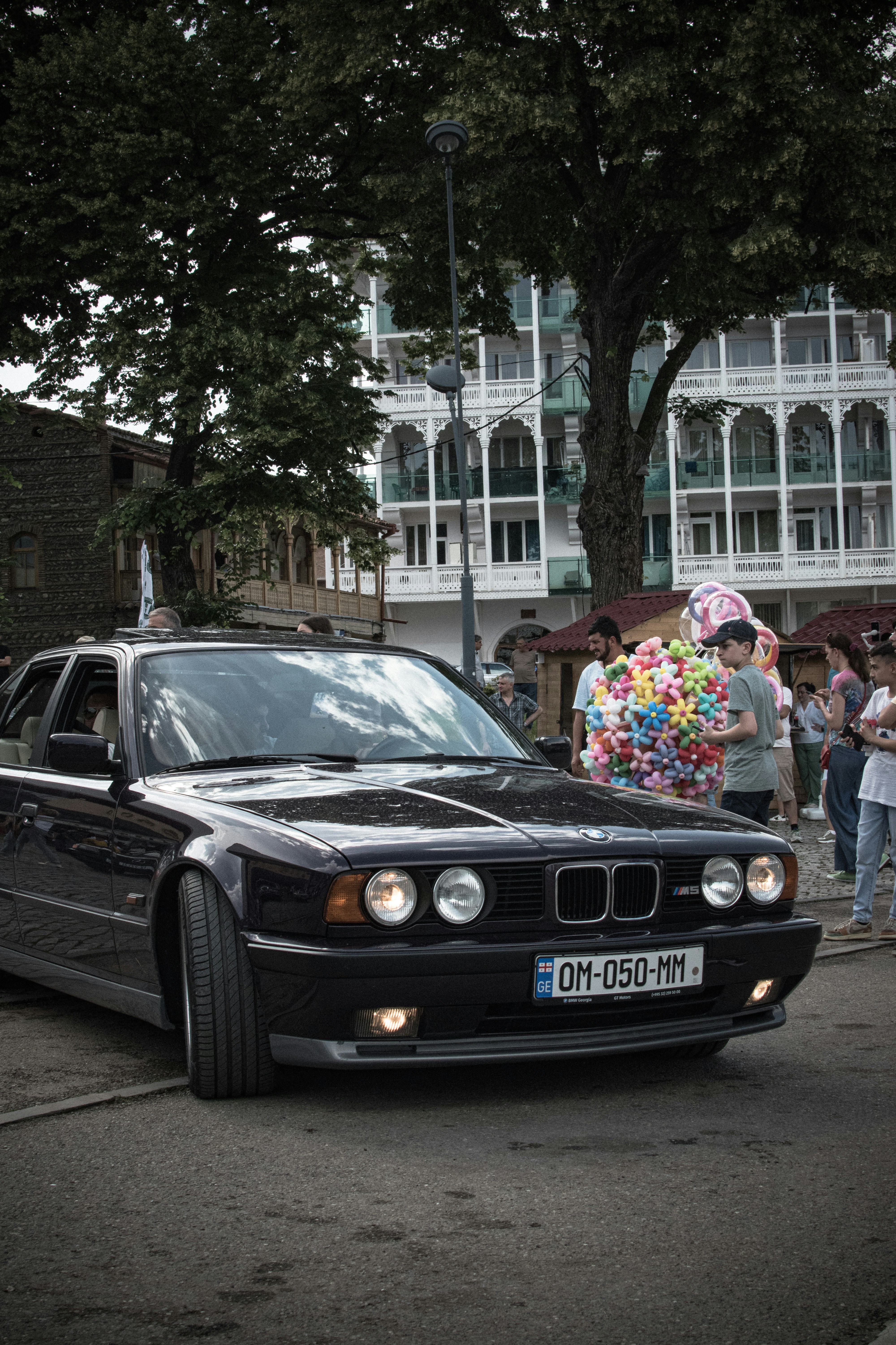 Black BMW in the Foreground, Crowds and Balloons Framing a City Street