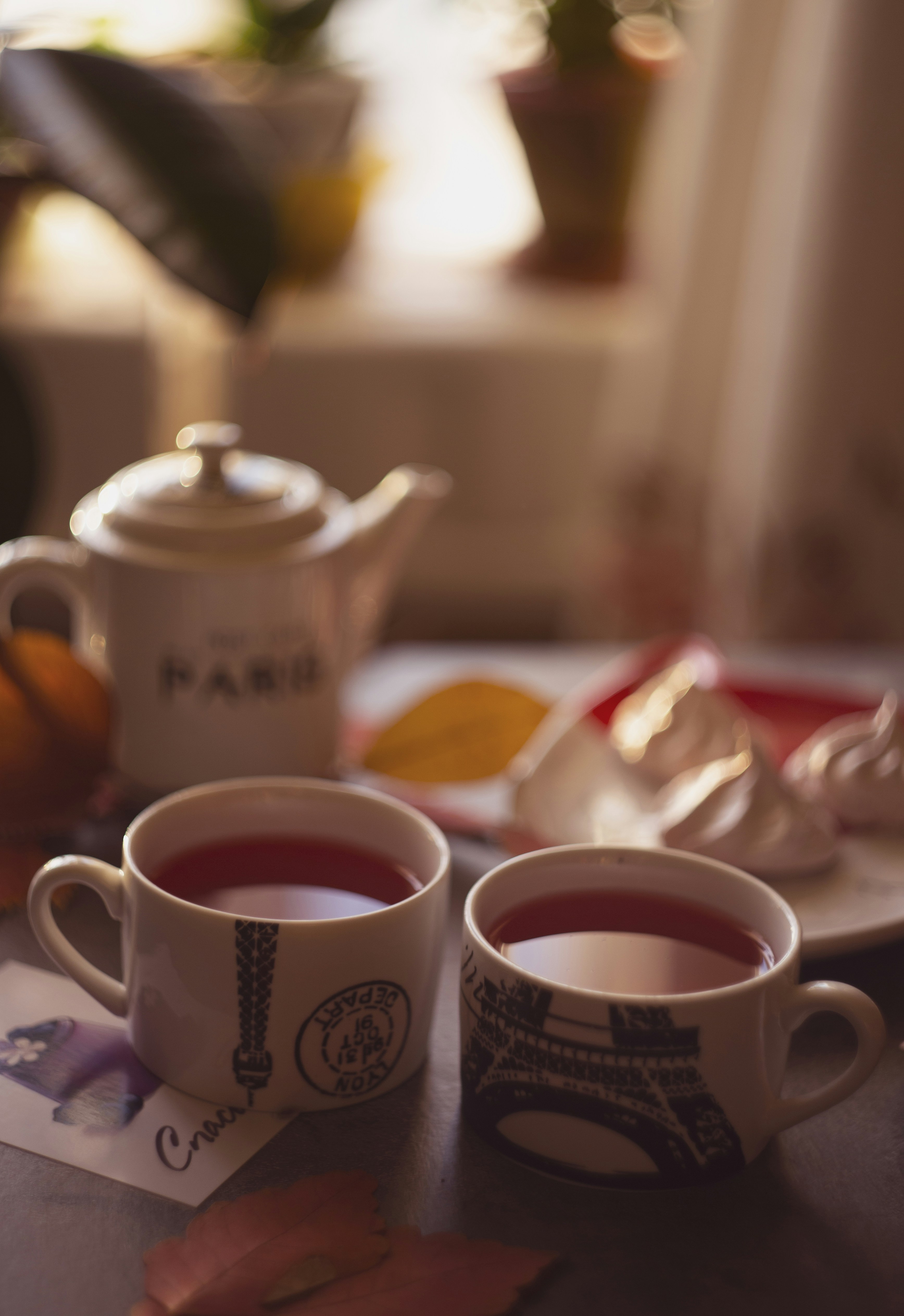 Tea-time in warm glow: two patterned cups foreground a teapot and card on a sunlit kitchen surface
