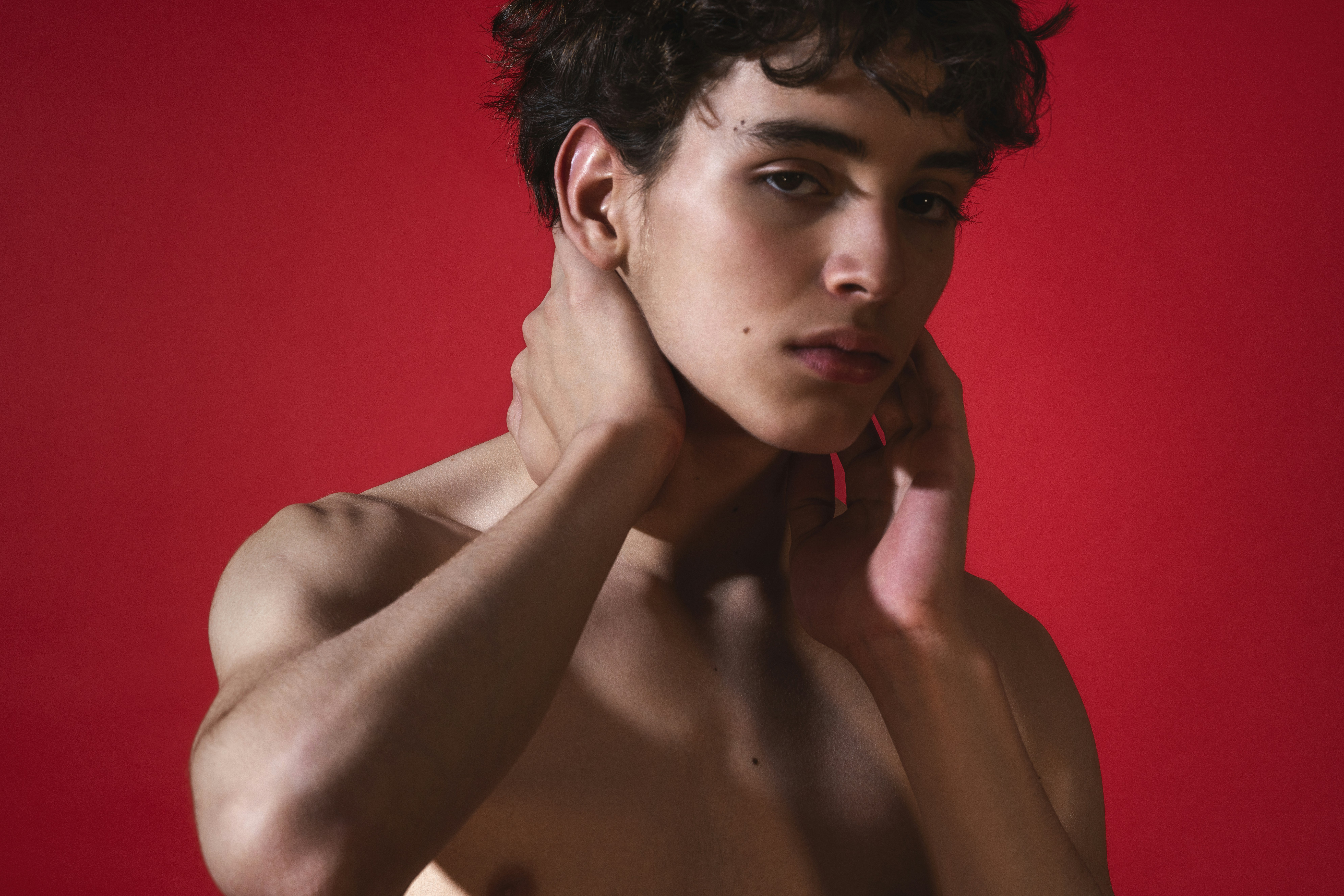 A young man poses against a vibrant red background, conveying a sense of introspection and vulnerability. The lighting accentuates his features and the mood of the scene.