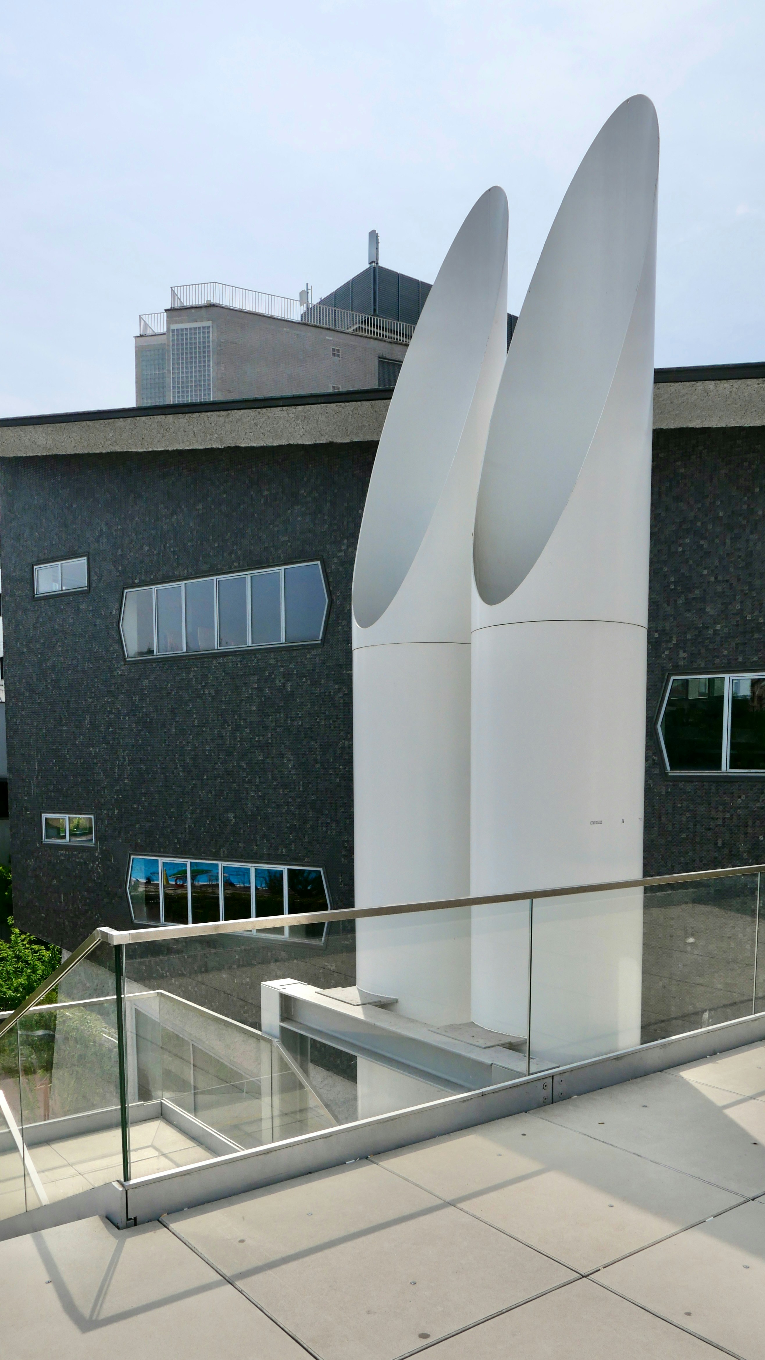 Modern architectural installation featuring two large, curved white structures against a textured gray building backdrop. The composition emphasizes sleek lines and contemporary design.