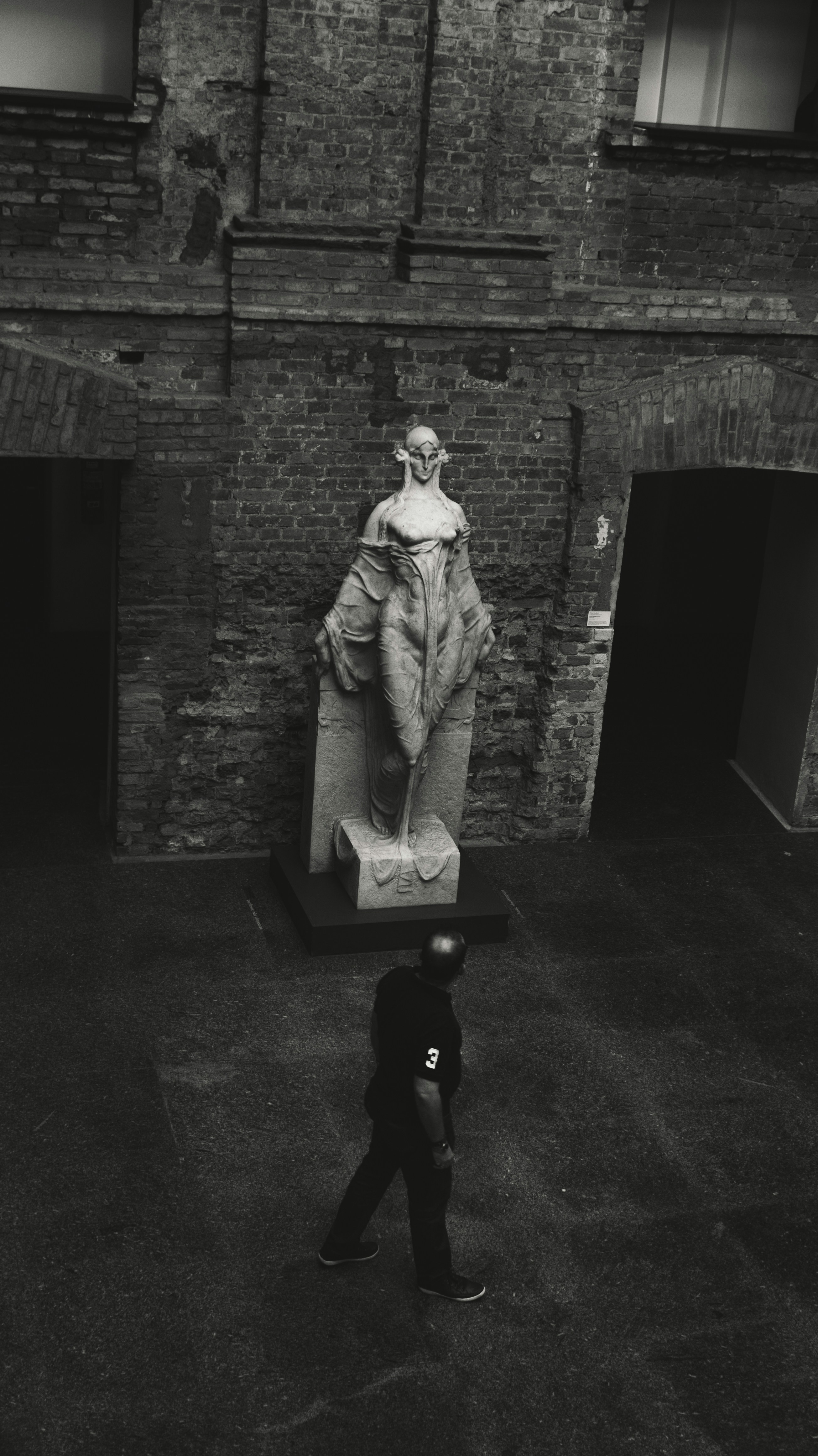 Monochrome Sentinel: A lone walker beside a stone statue in a brick courtyard