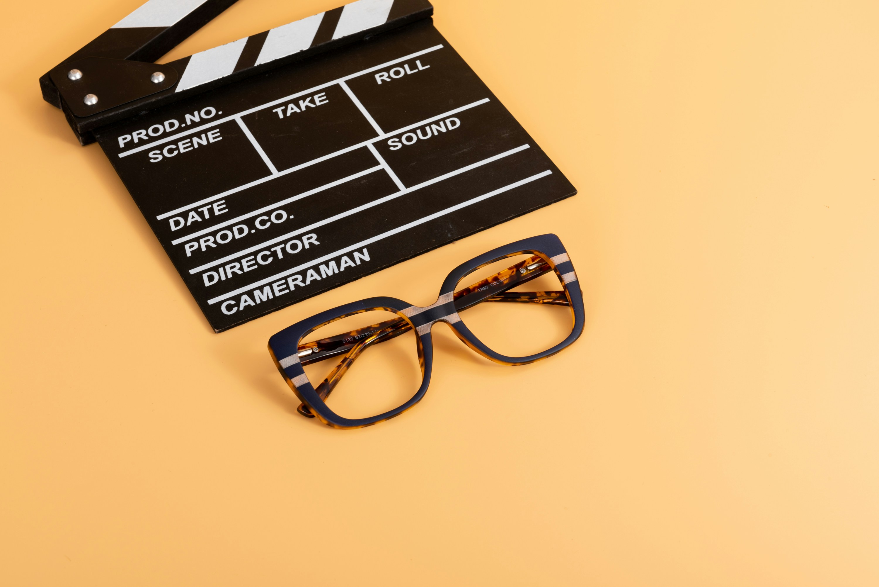 A pair of stylish, tortoiseshell and navy blue eyeglasses lies next to a classic movie clapperboard on a vibrant orange background. The sleek design of the frames, combined with the film industry prop, evokes a sense of cinematic flair and artistic sophistication, ideal for fashion-forward creatives. | A movie clapper, glasses and a pair of reading glasses on a yellow background