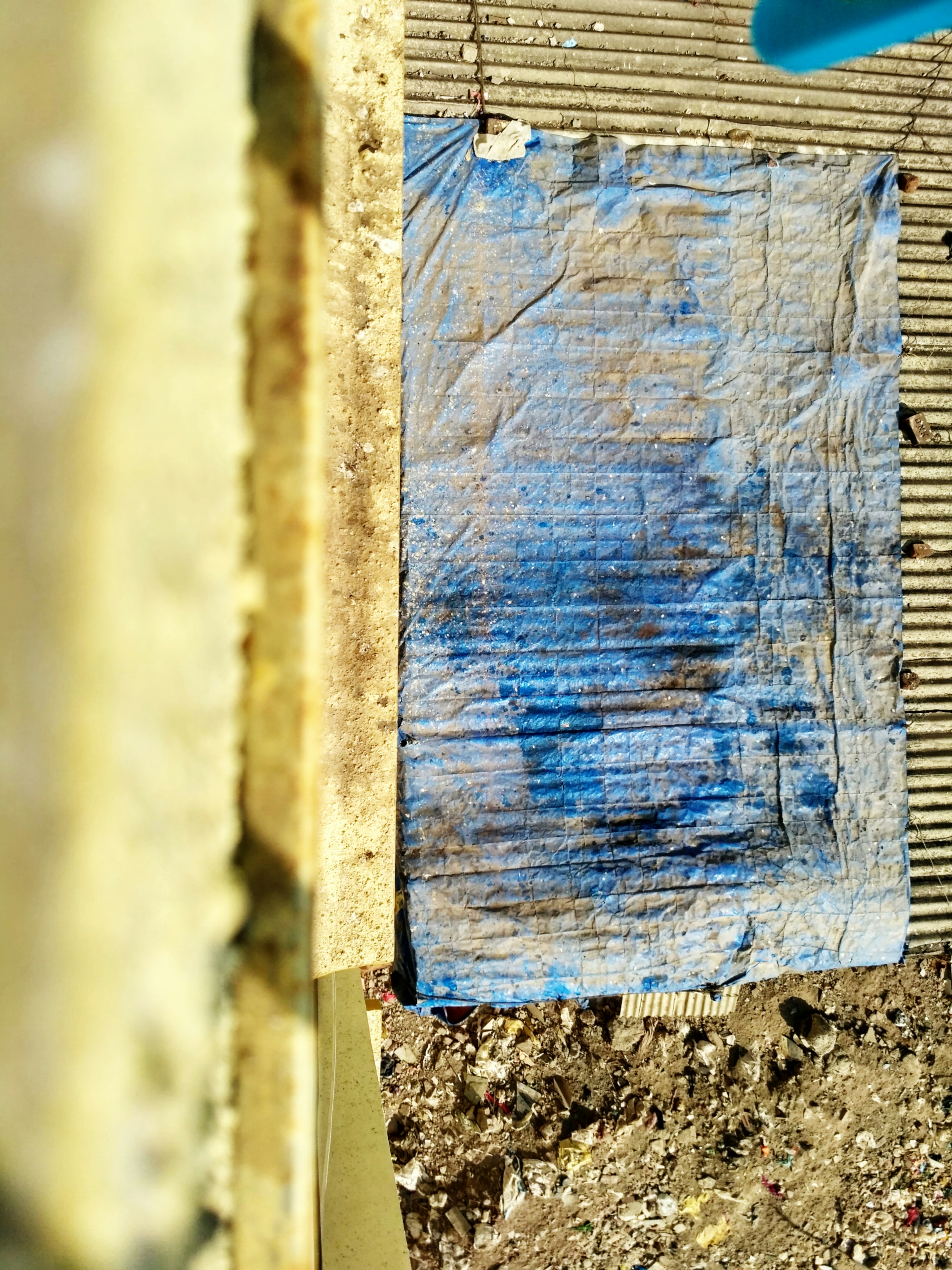 Blue tarpaulin lies on a dirt-covered construction site, framed by a yellow wall and wooden planks. The scene highlights rough ground textures and weathered fabric.