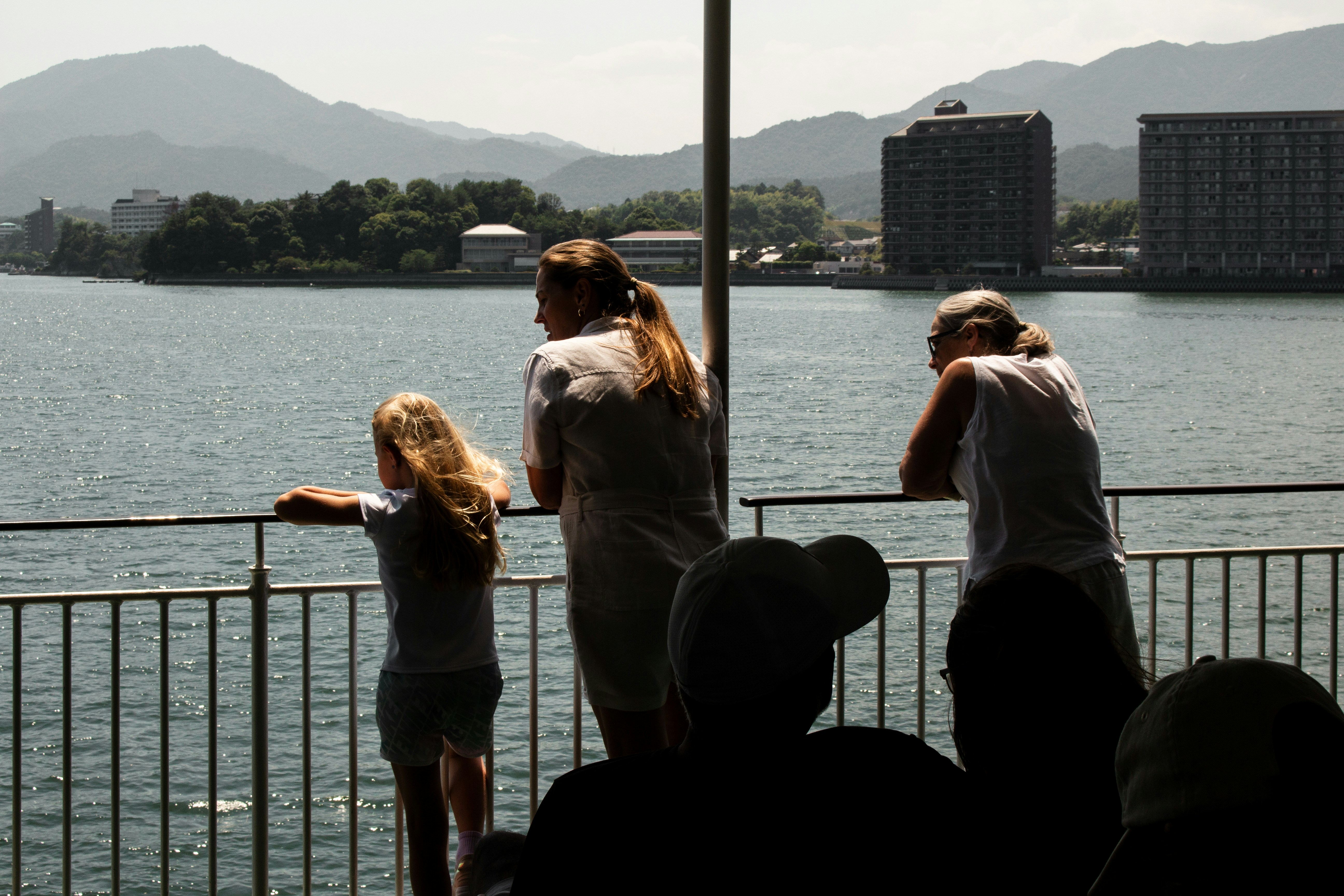 Three people standing on a ferry railing, gazing across the water with distant mountains in the background.