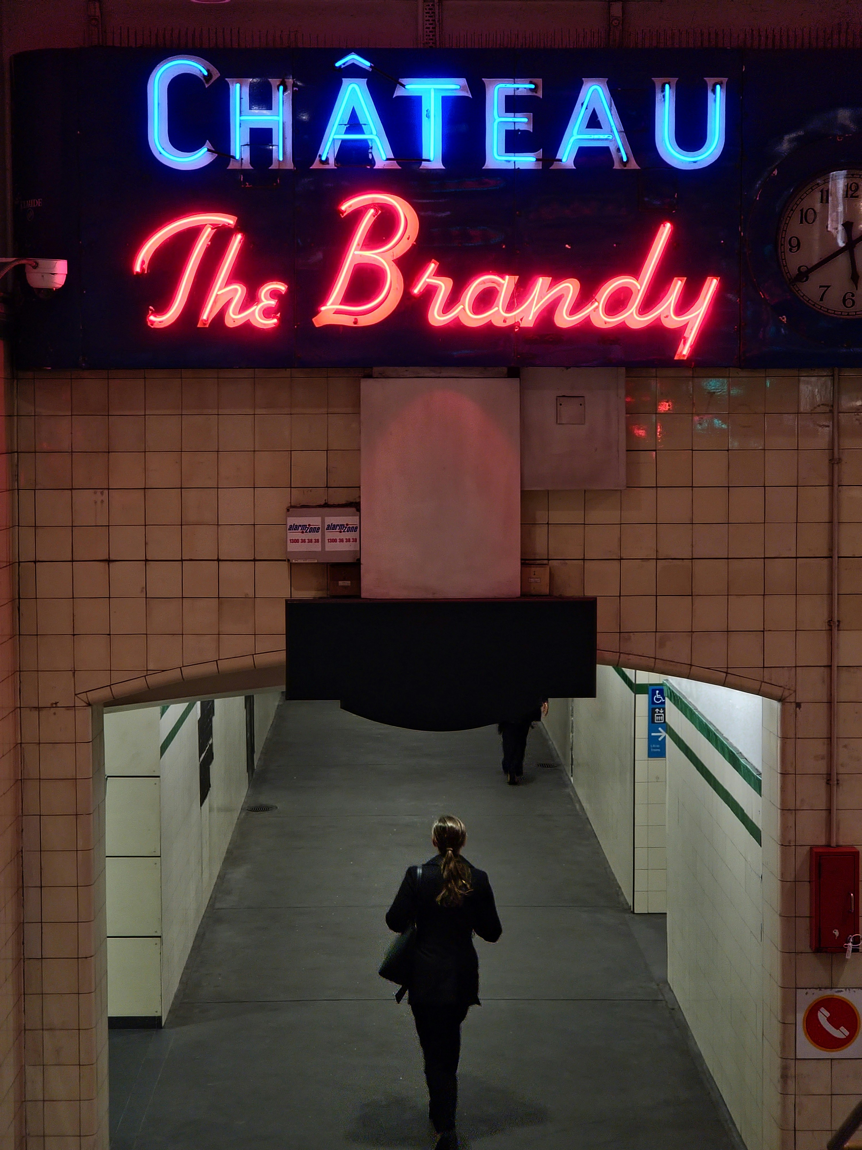 Neon Threshold Under Château Brandy Sign: A lone walker in the subway corridor