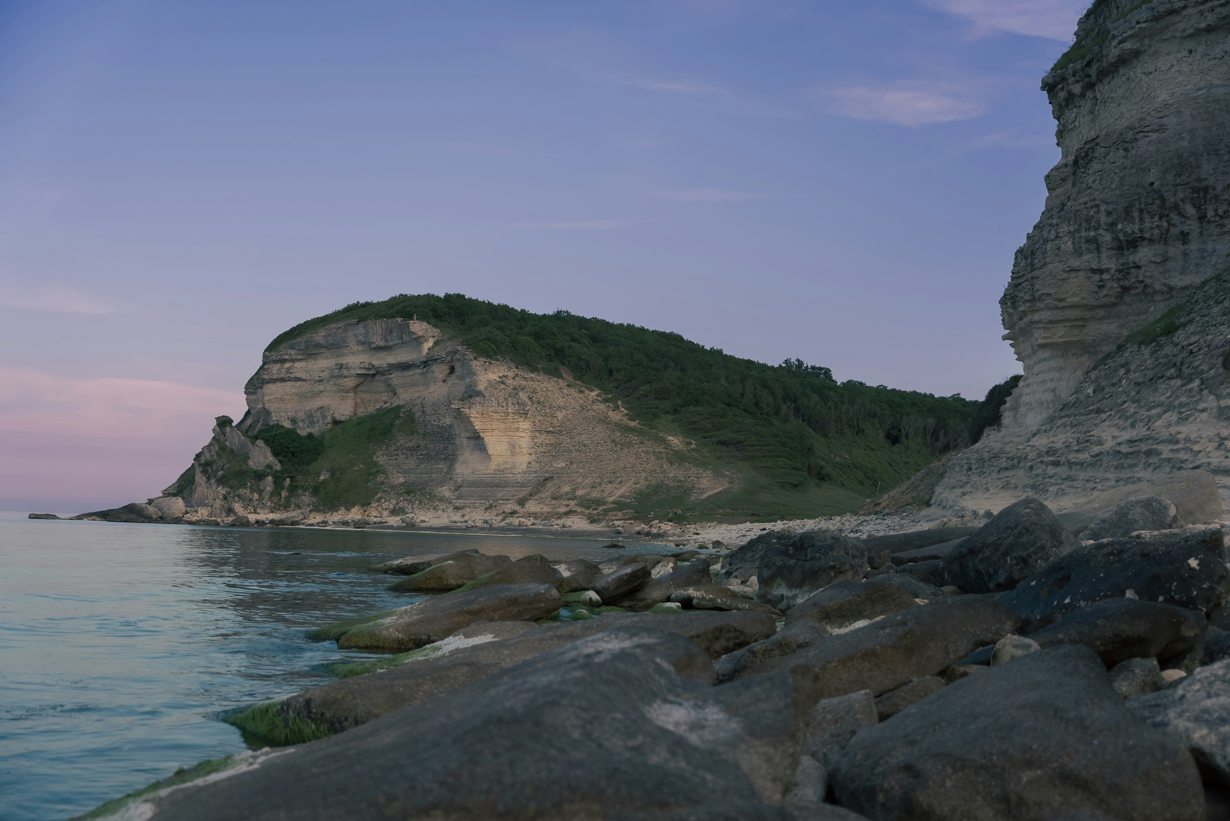 A photograph of a green-topped limestone headland extending into calm water, with a rocky foreground. Soft pastel sky and gentle waves complete this tranquil coastal scene.