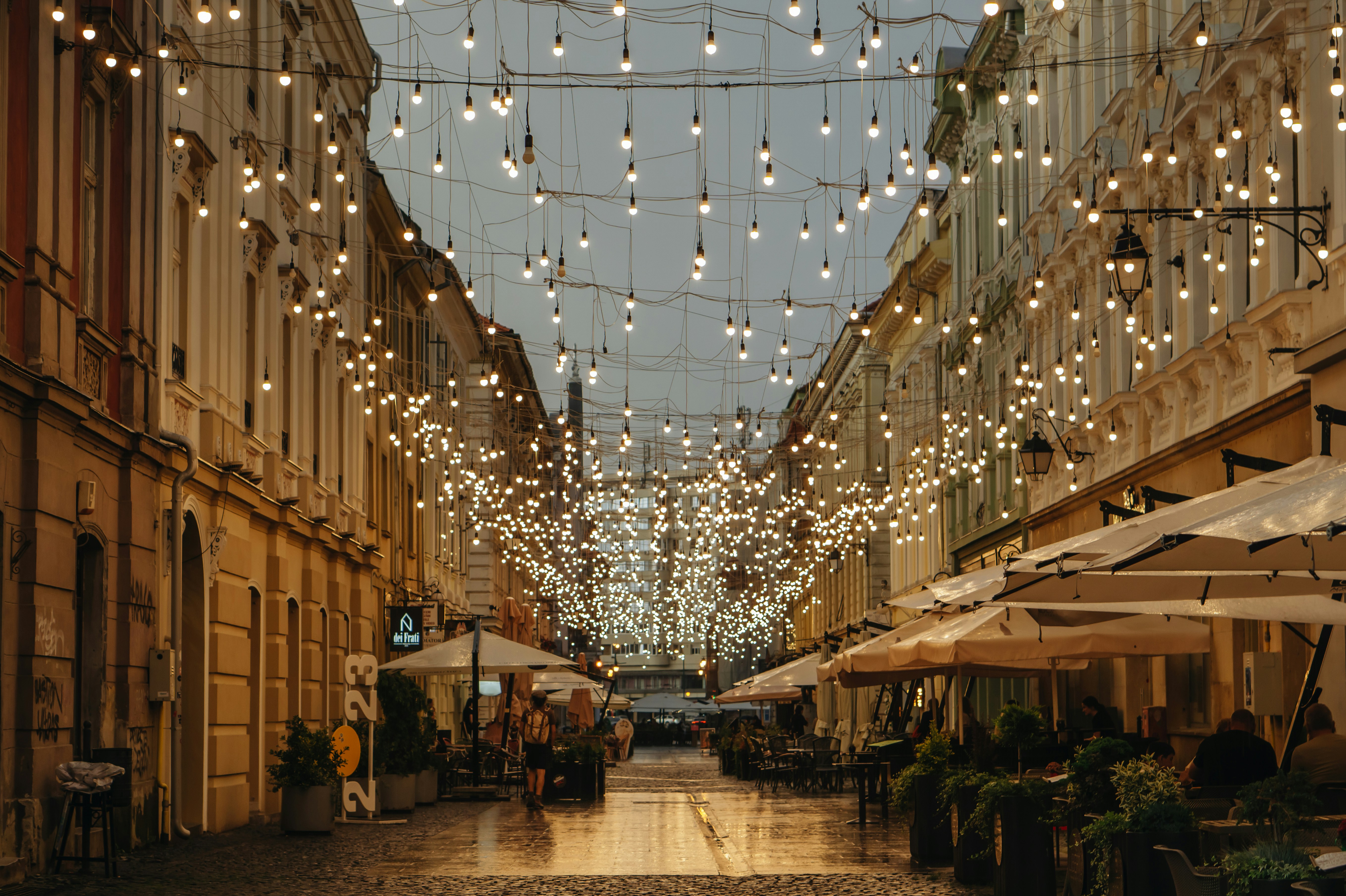 String lights illuminate a narrow street lined with outdoor cafes and historic buildings at dusk.