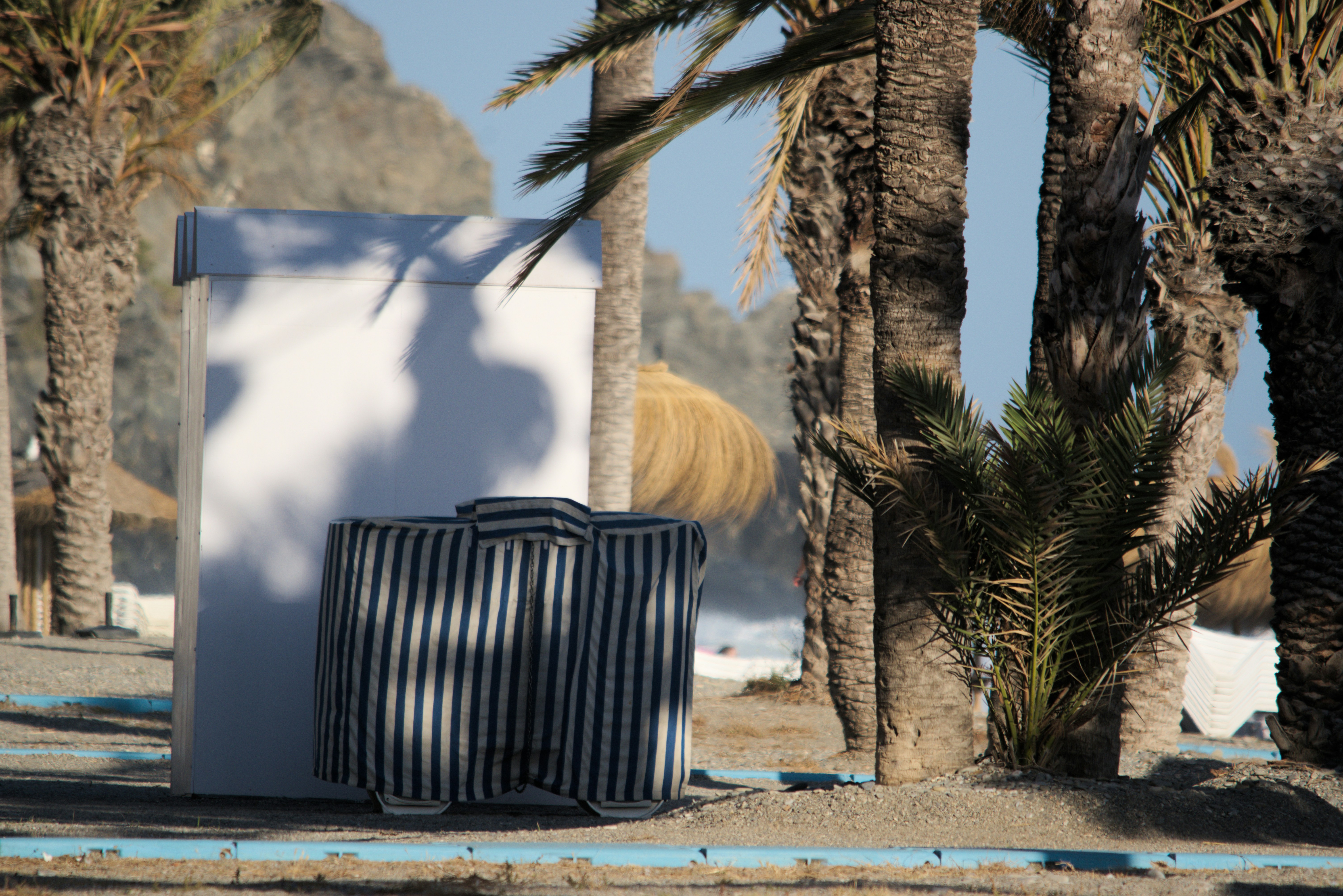 Shadowed seaside scene: striped bag under palm treetops near a white cabana