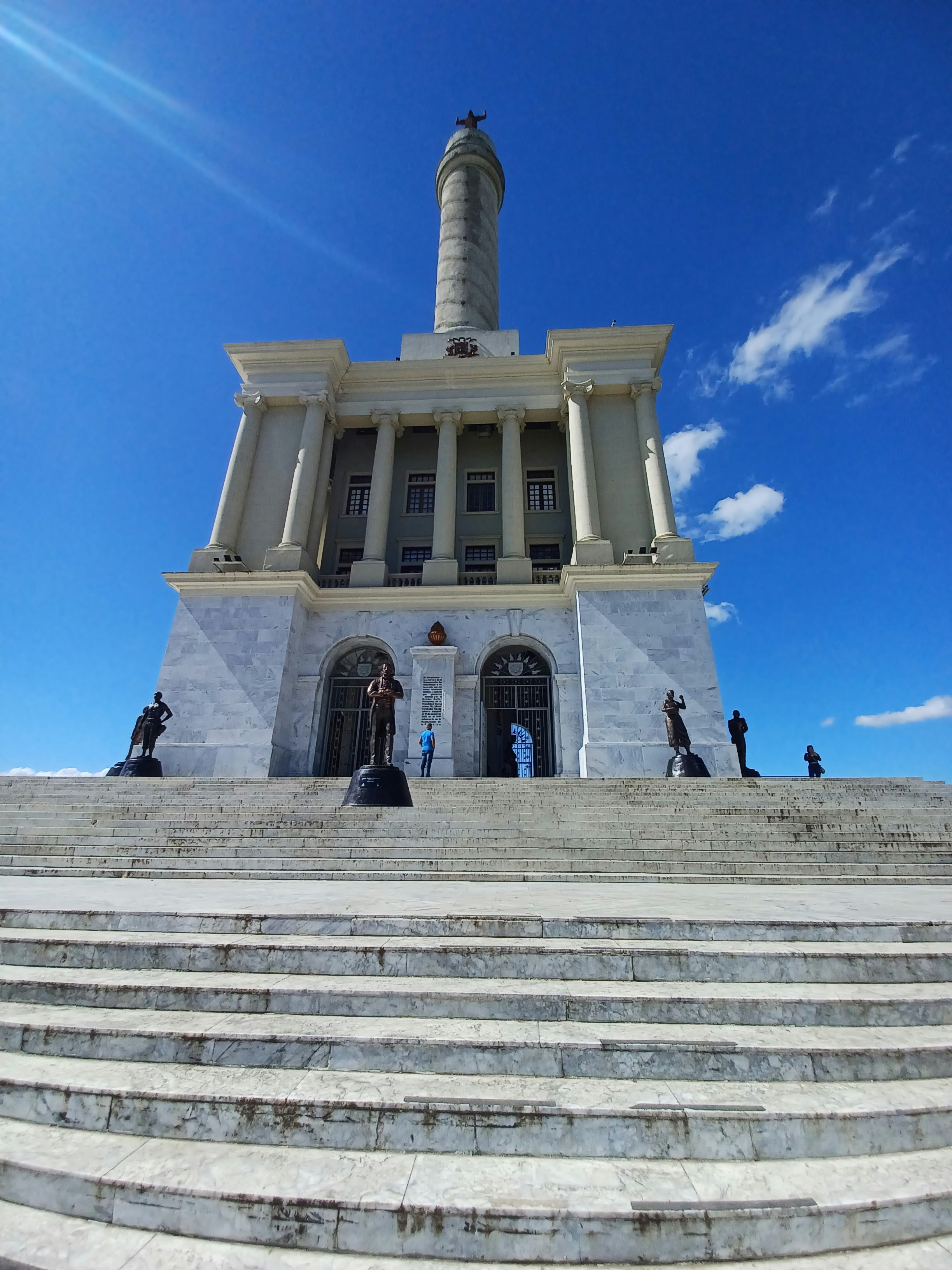 White marble monument with a tall central column and classical façade rises above a broad staircase, flanked by statues. The scene is set beneath a clear blue sky.