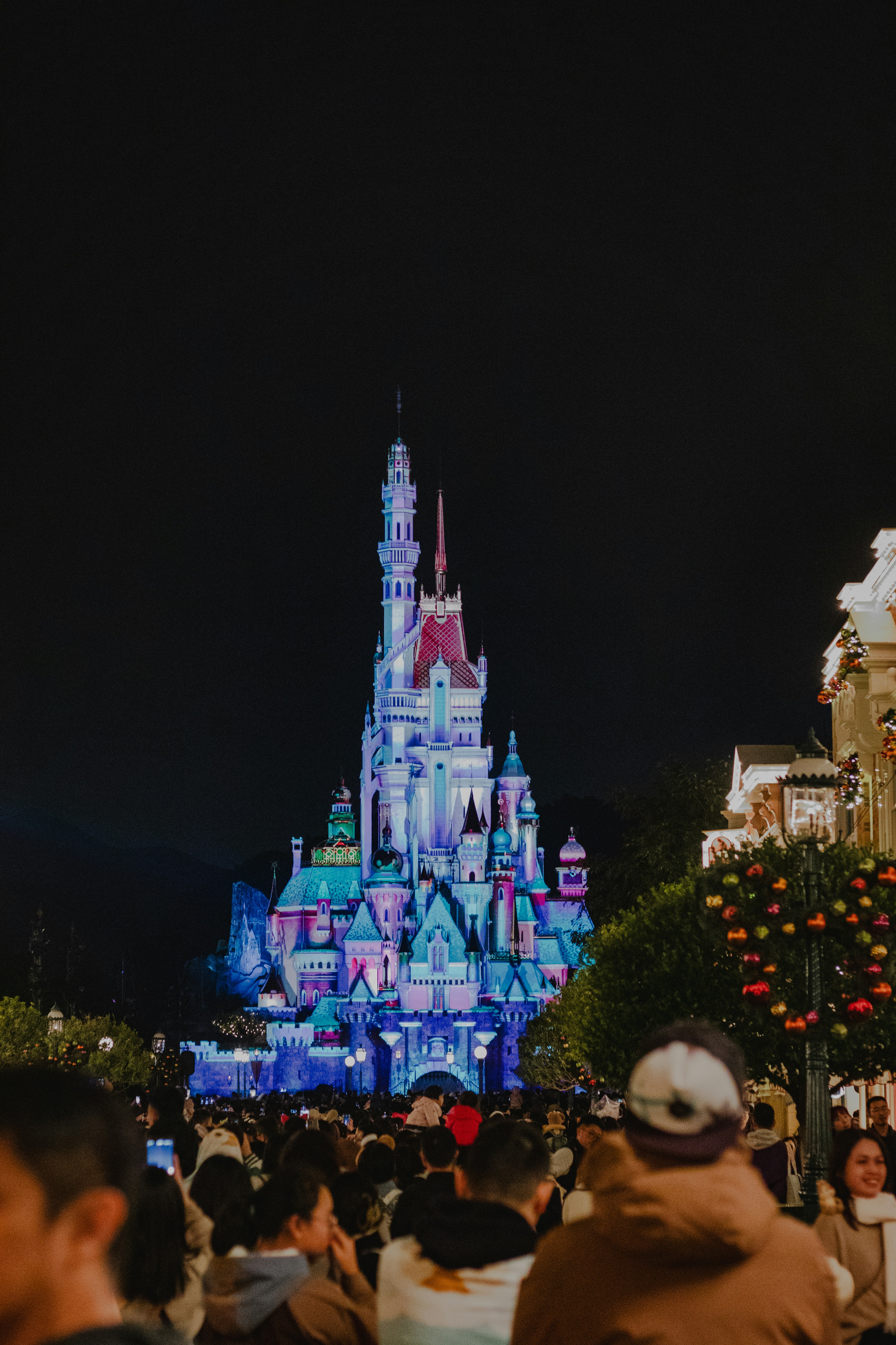 Vibrantly lit castle surrounded by a crowd during a nighttime event, showcasing colorful lights and architectural details.