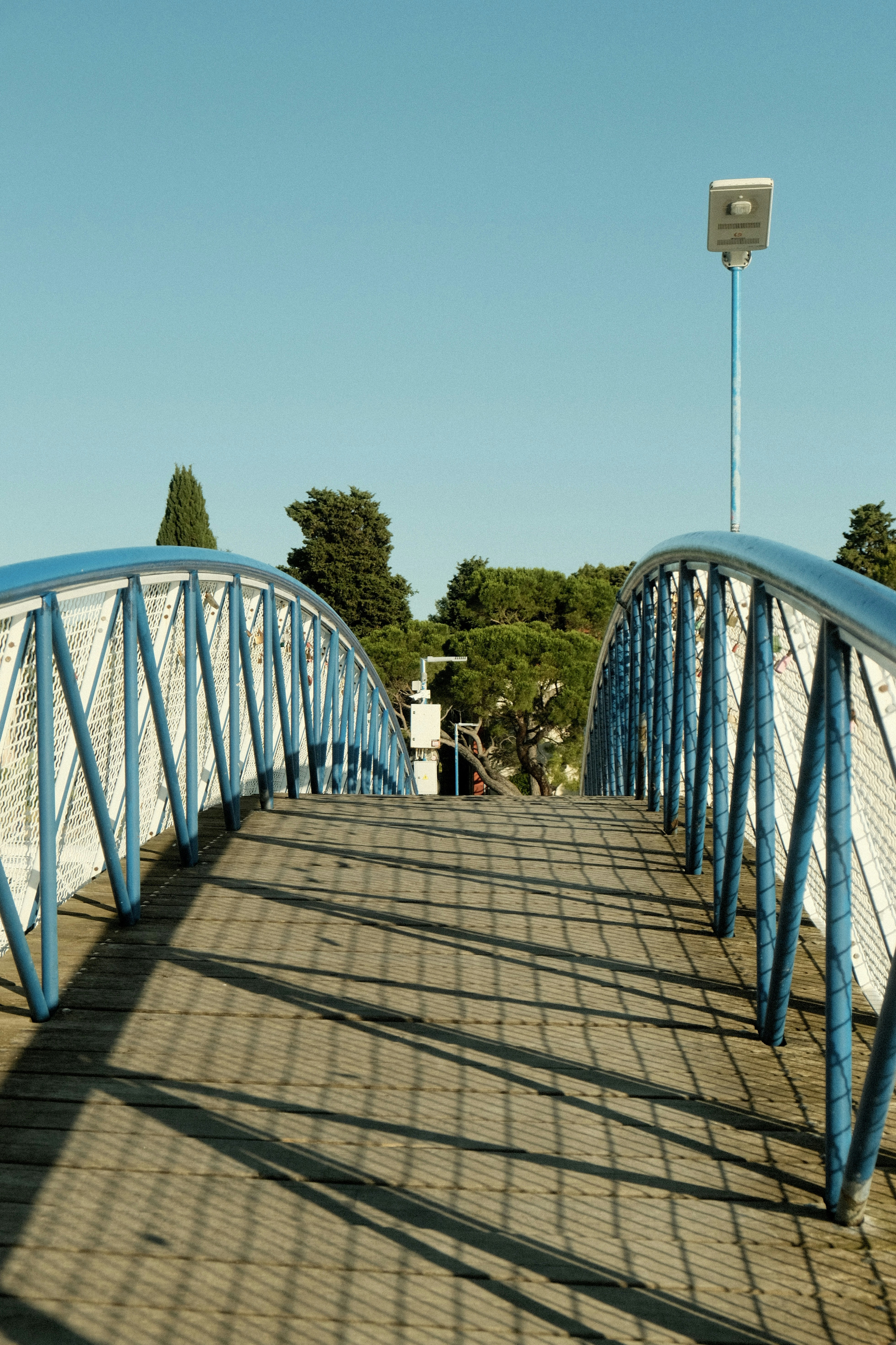 Blue Arches and Long Shadows: A Pedestrian Bridge Toward Sunlit Greenery