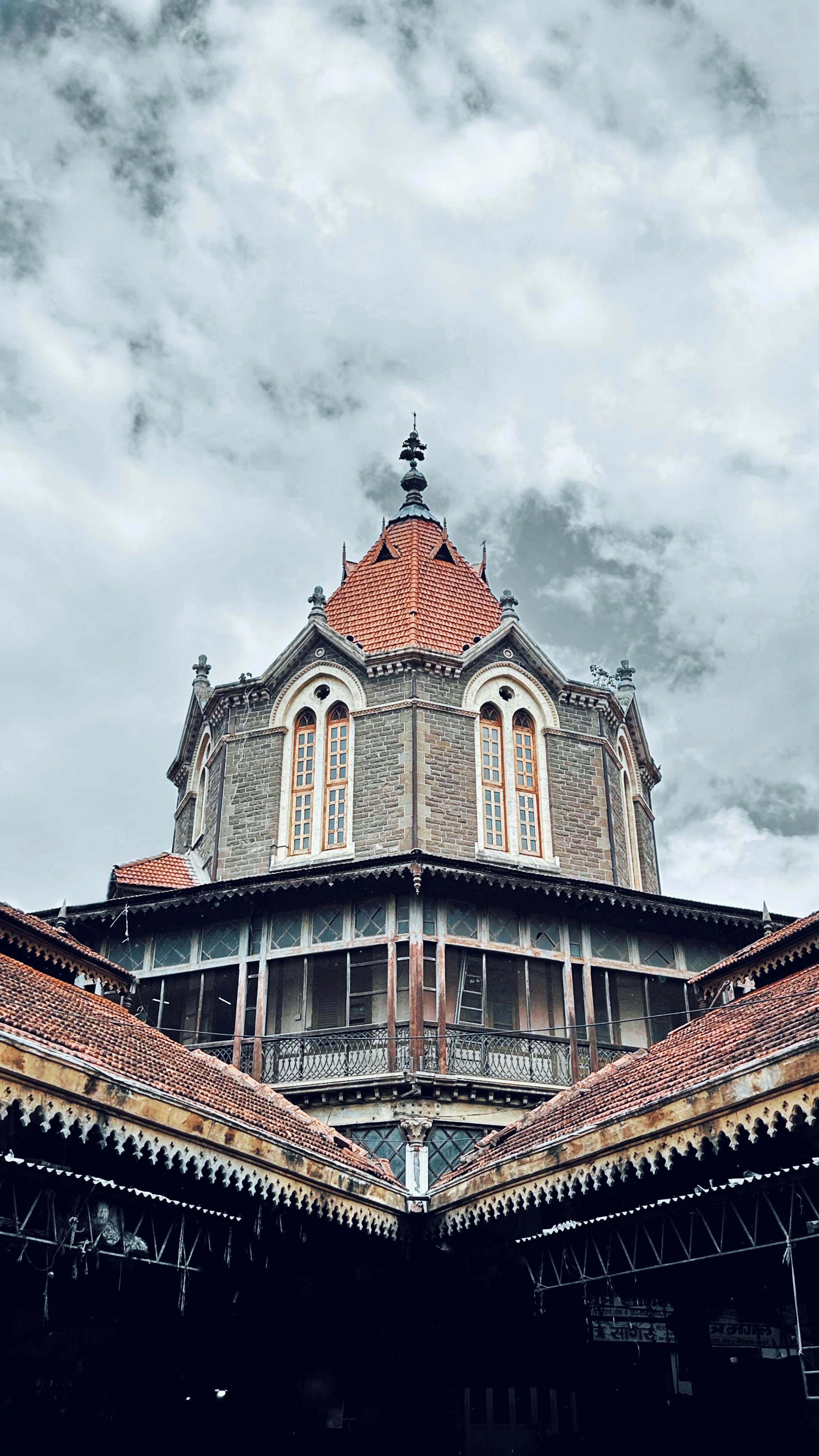 Neo-Gothic tower with a terracotta red roof dominates the center, rising above a symmetrical courtyard. This photograph emphasizes architectural detail against a dramatic, cloudy sky.
