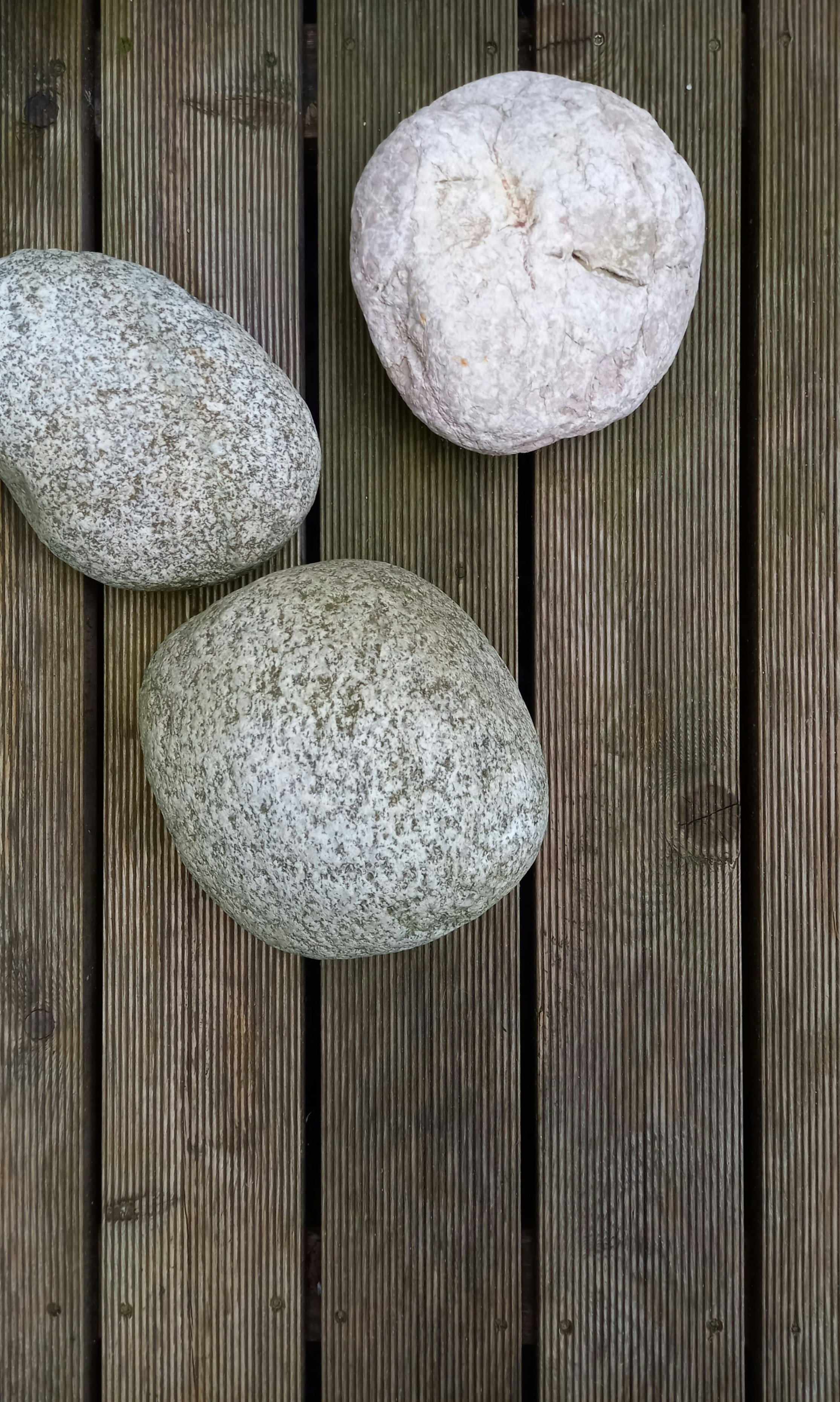 Three rounded pebbles in muted greys rest on weathered wooden deck planks, forming a simple still-life composition.