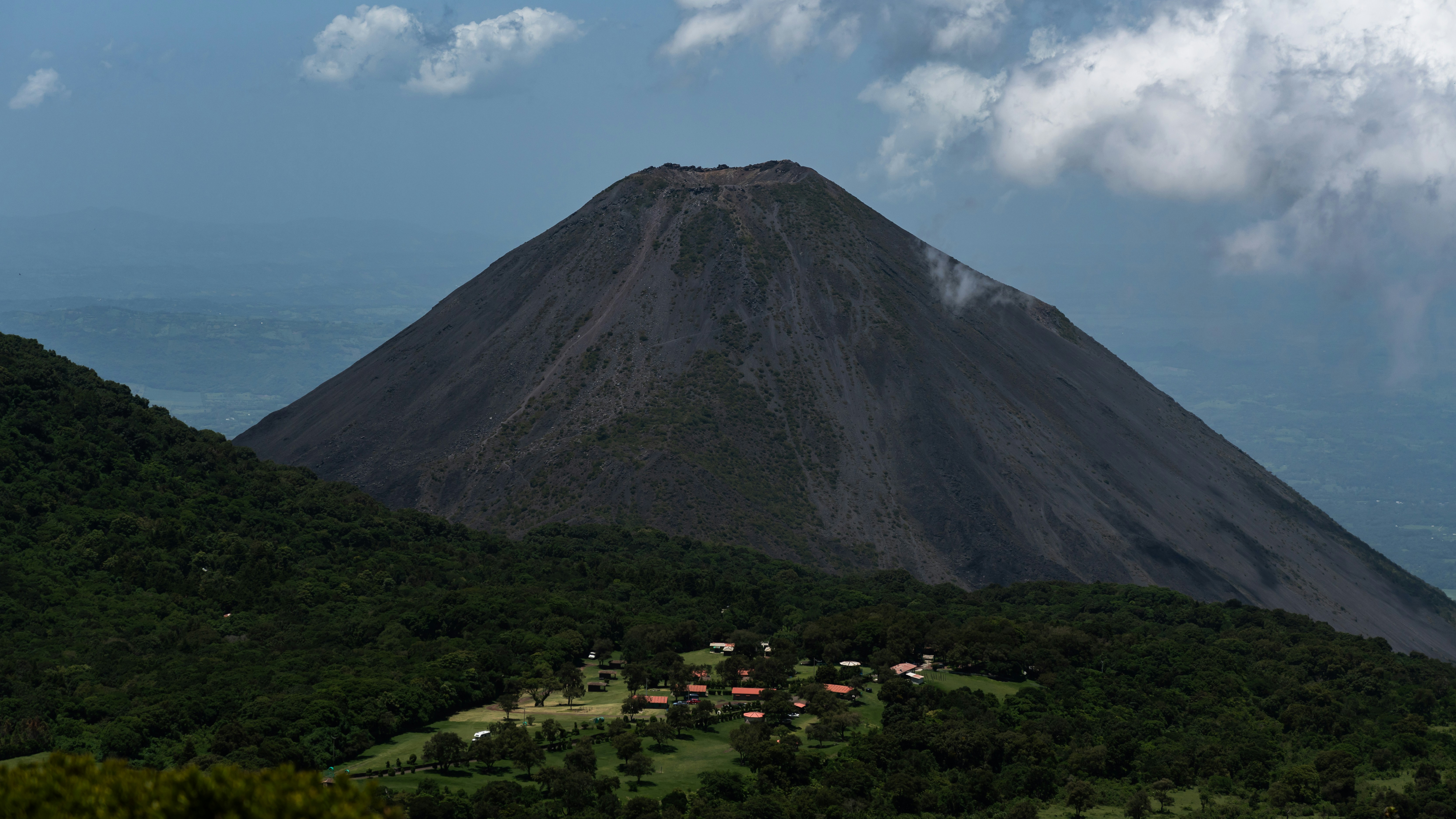 Volcanic mountain rising above lush green hills with a cluster of small buildings at its base under a partly cloudy sky.
