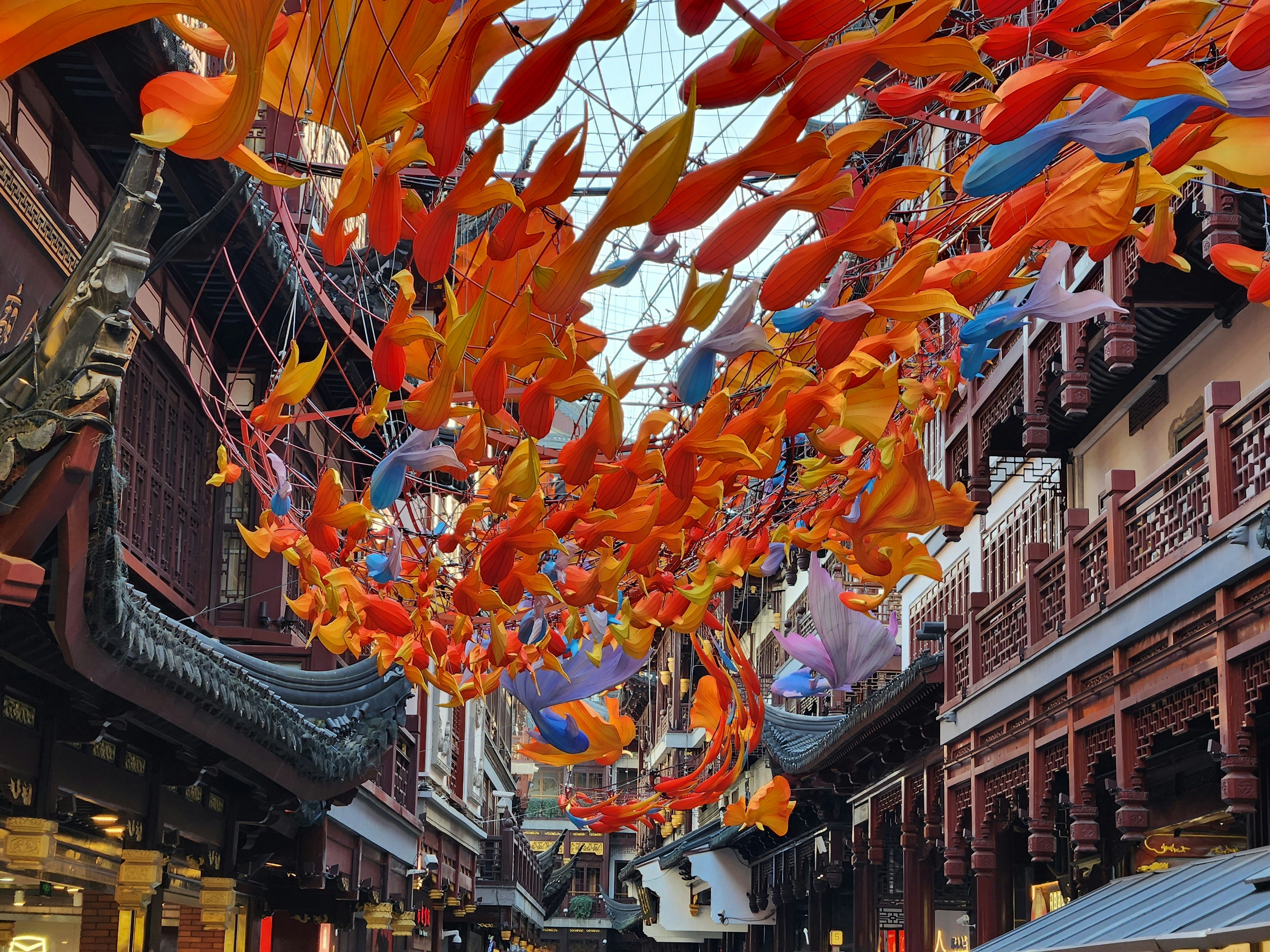 A narrow alley flanked by traditional wooden buildings is capped by a suspended canopy of orange and multicolored fabric, forming a vivid tunnel. The shot emphasizes perspective and color as the canopy leads the eye toward the distant vanishing point.