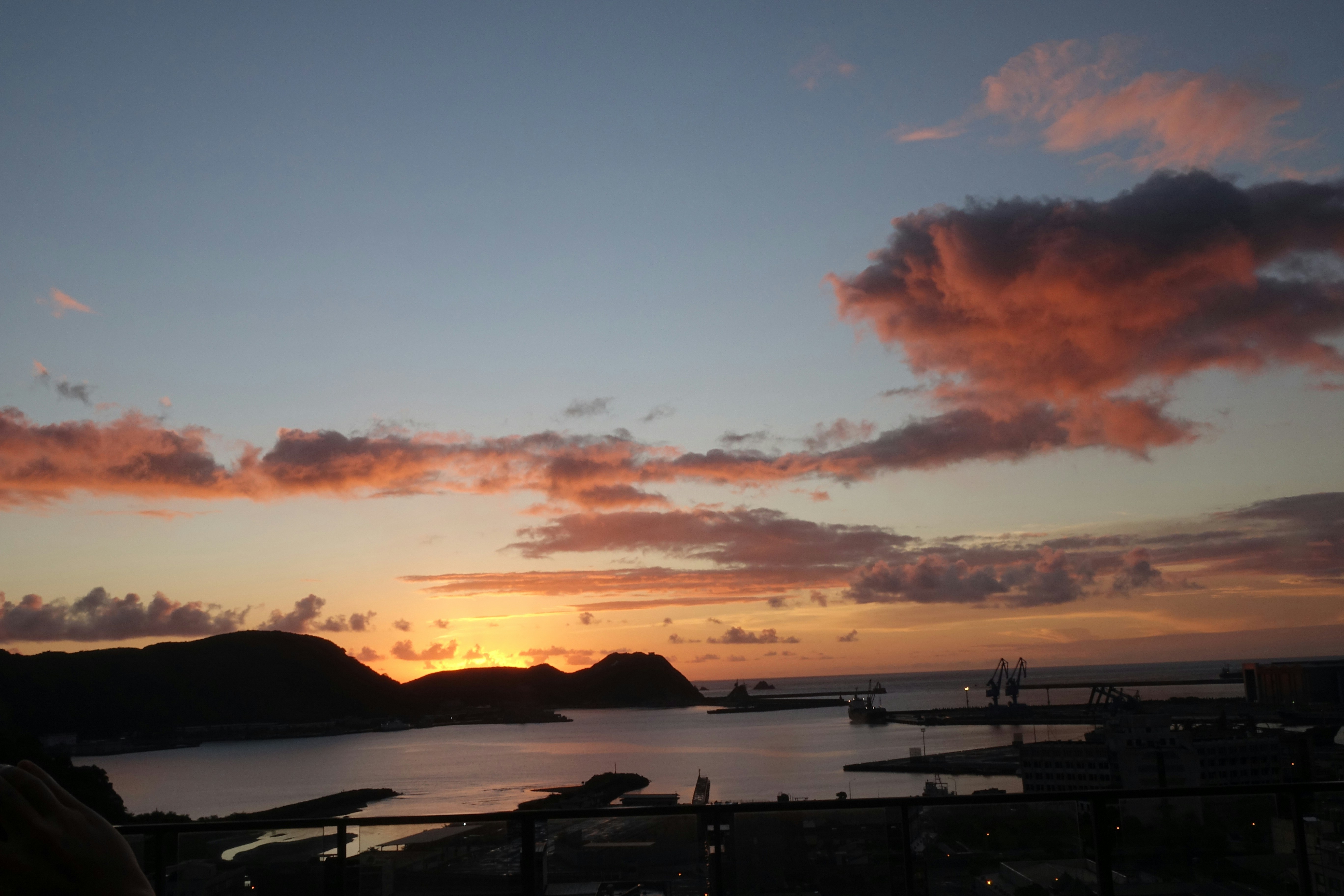 Vivid sunset clouds reflect over a serene harbor and silhouetted hills.